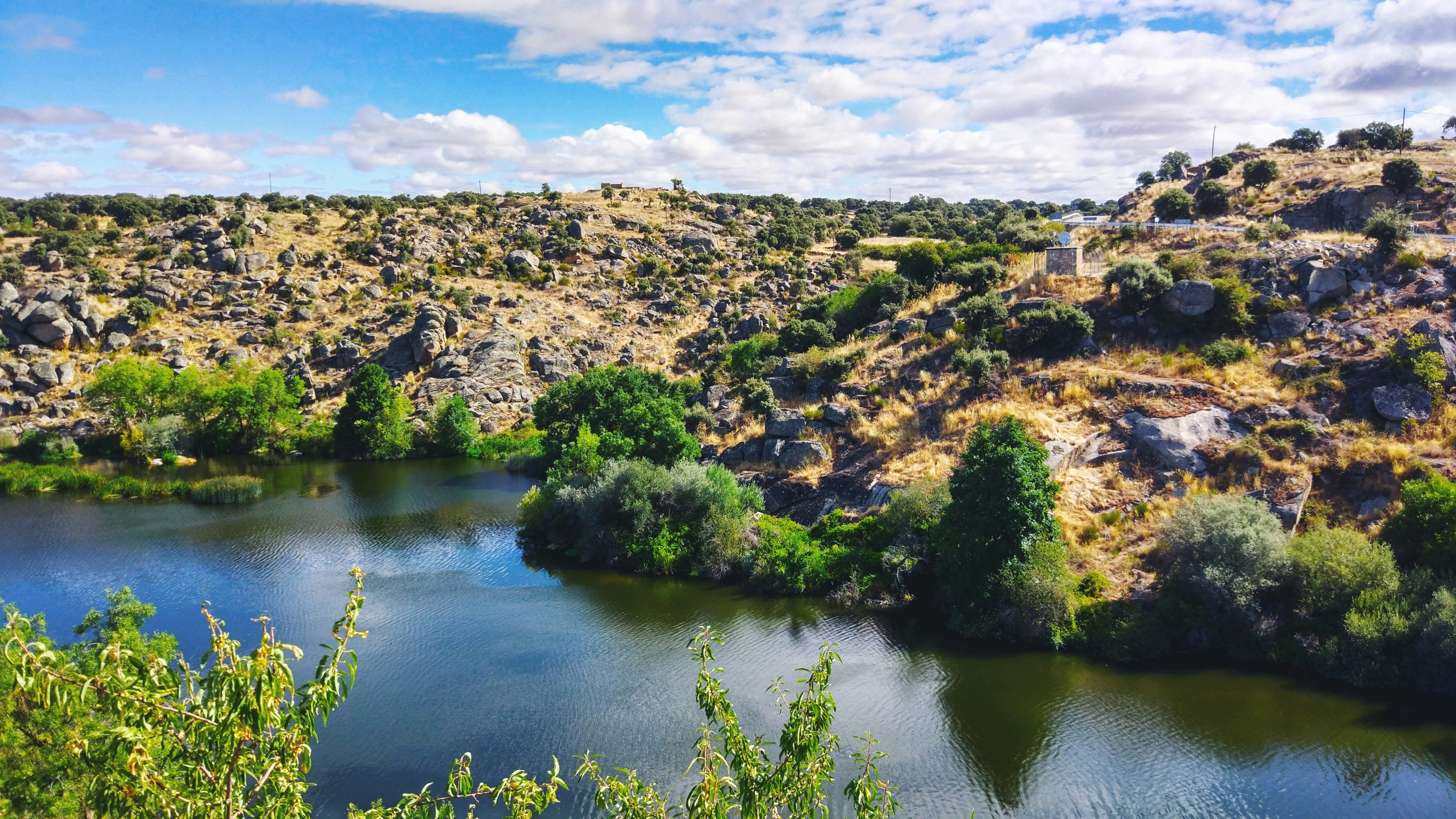 Calm river reflecting rocky hills and scattered trees under a partly cloudy sky.
