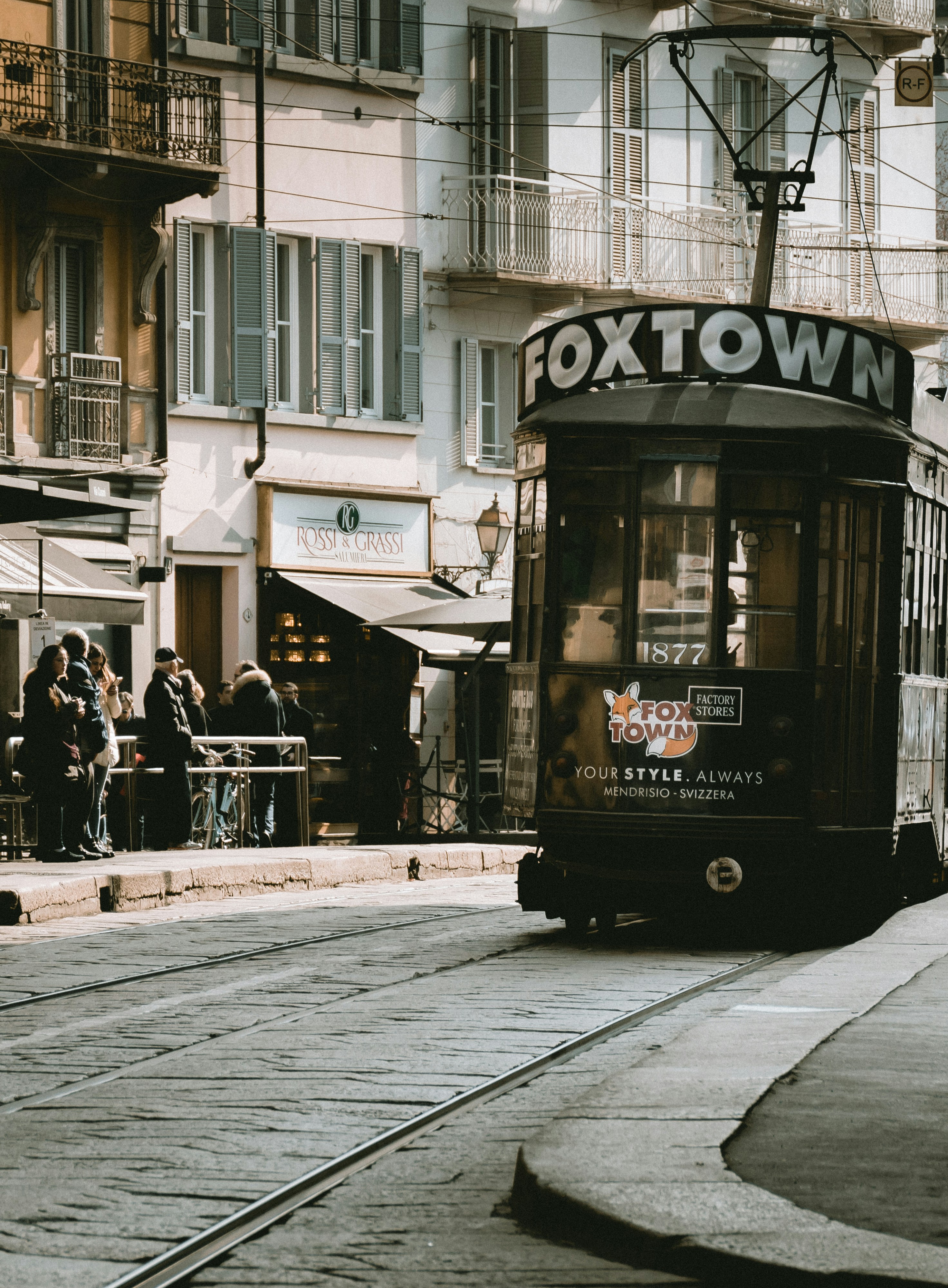 A vintage tram labeled 'FOXTOWN' navigates a cobblestone street, surrounded by quaint shops and a gathering of people. The scene captures a moment of urban life in a historic setting.
