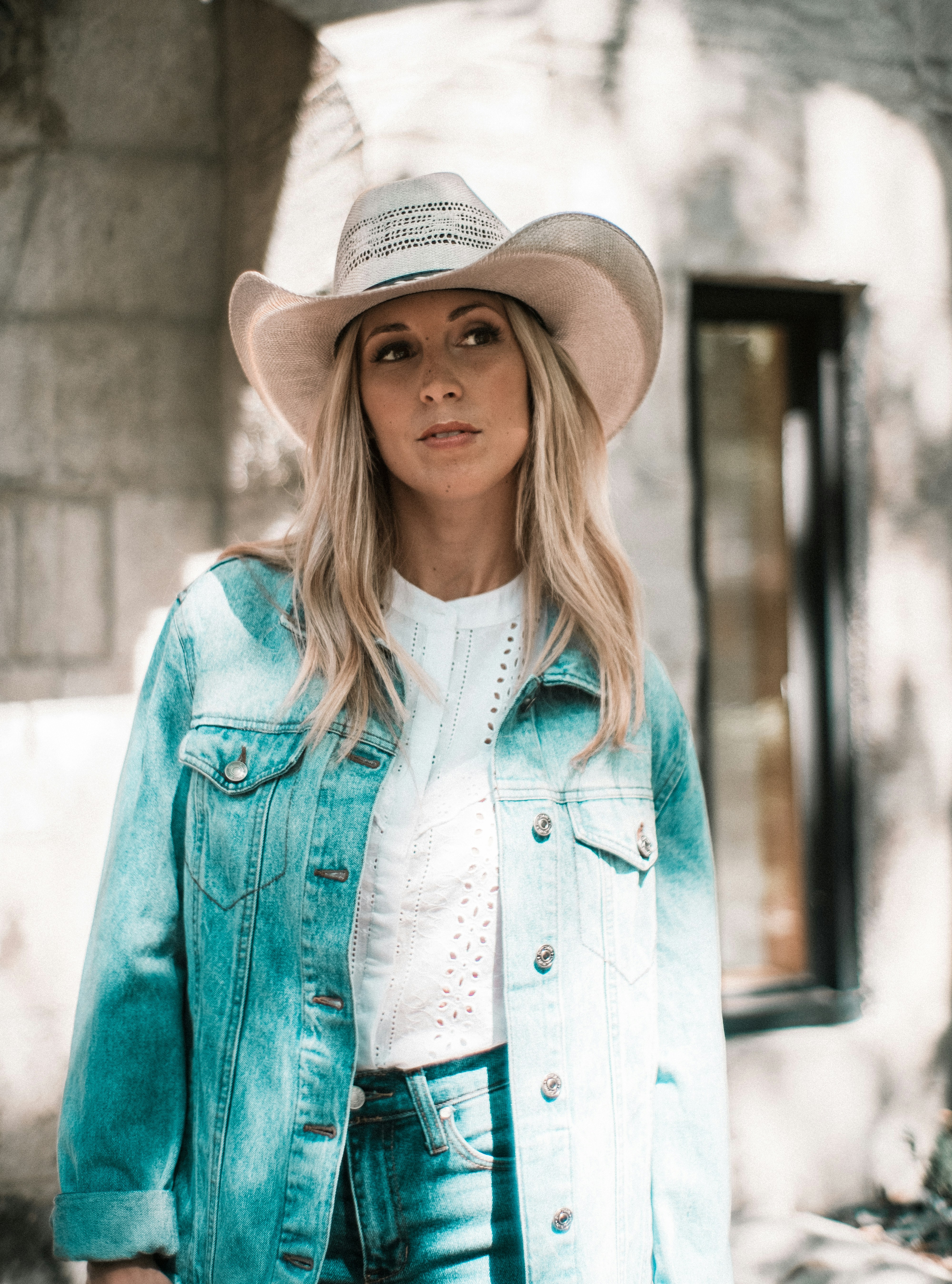 A woman in a denim jacket and wide-brimmed hat stands against a textured stone wall, embodying a blend of rustic and modern styles.