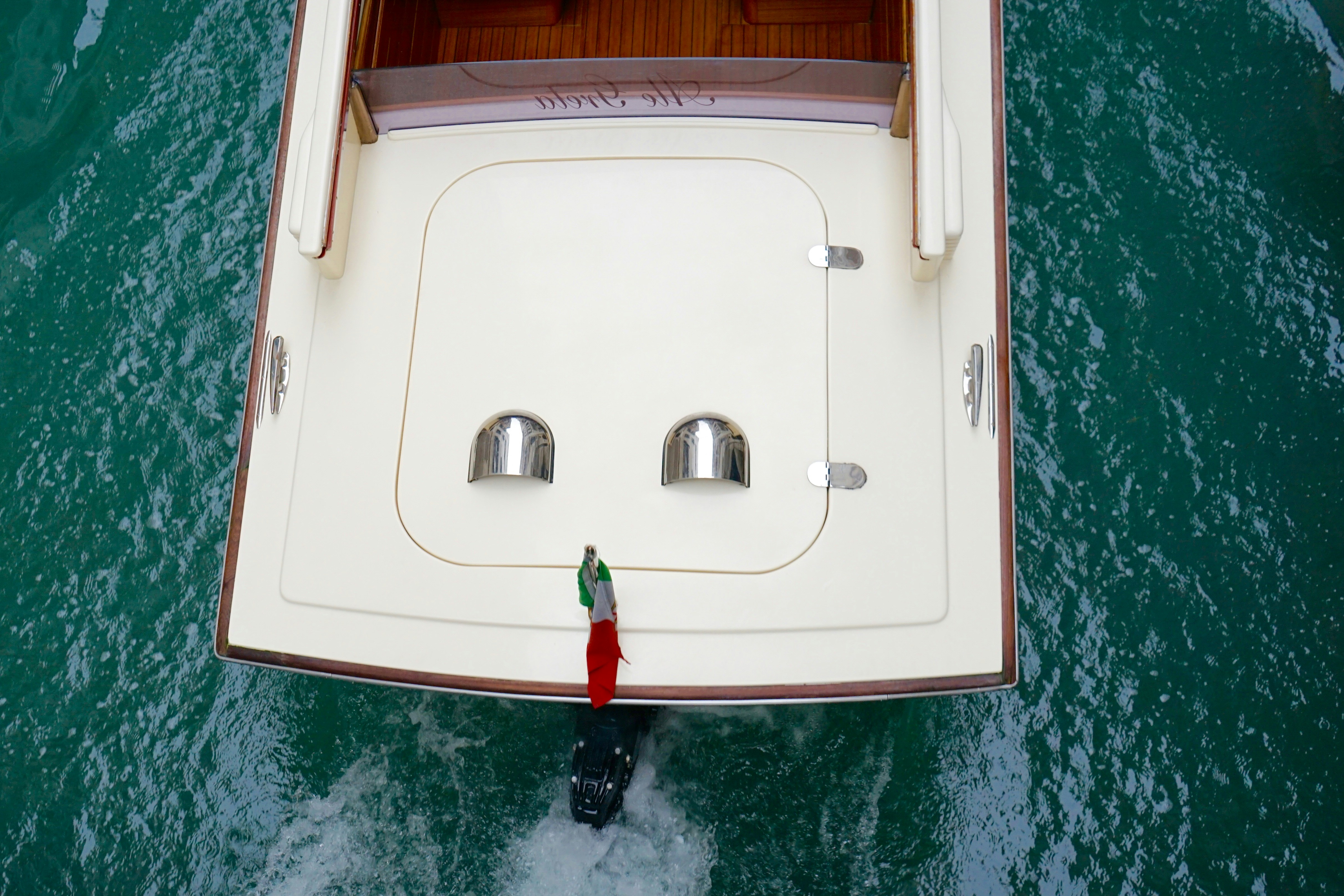 Aerial photograph of a person in green shirt and red pants standing at the stern of a white yacht deck, surrounded by turquoise water.