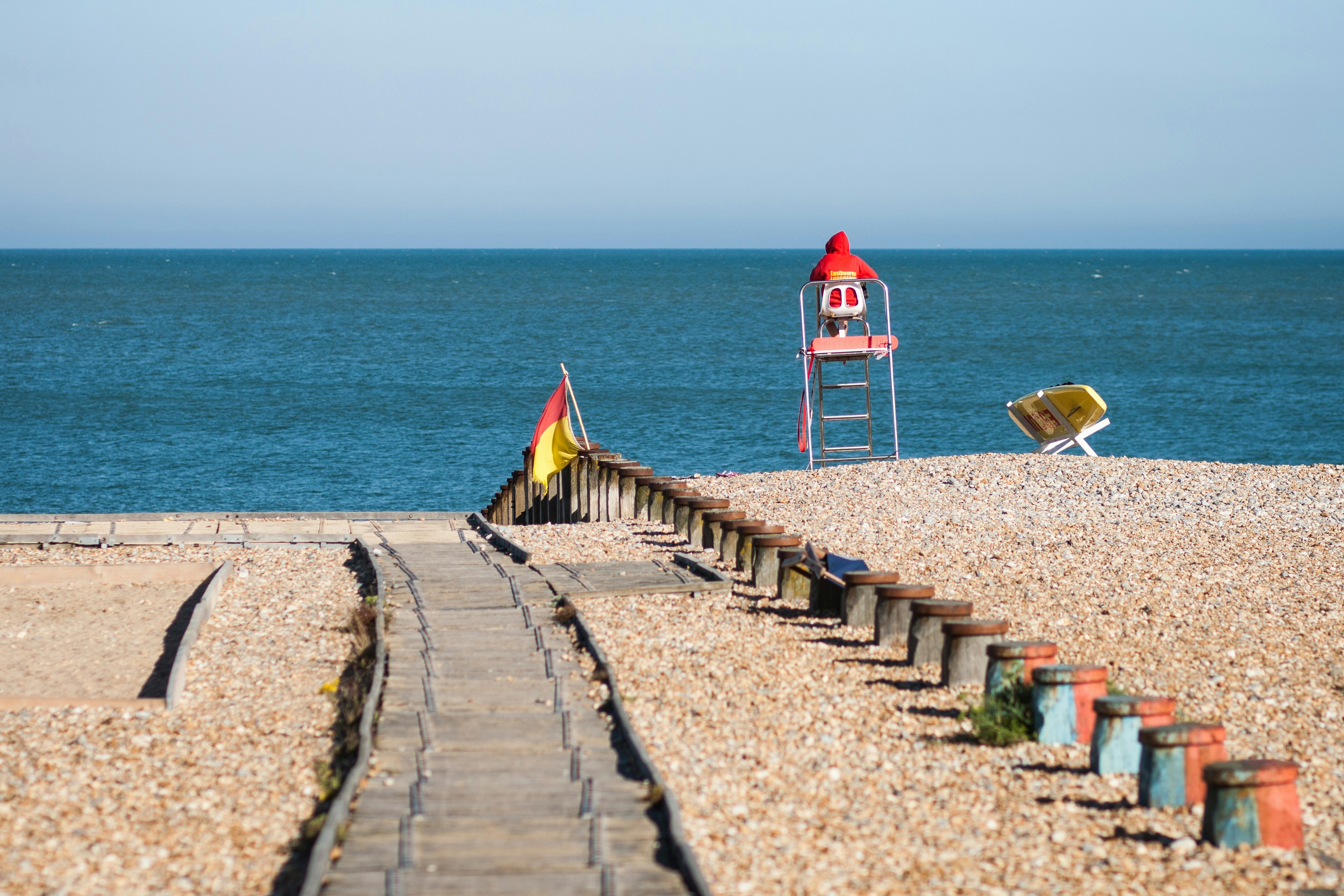 Wooden boardwalk leading to a pebbled beach with a lifeguard tower and calm blue sea.