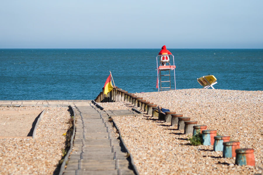 A lone lifeguard in a red jacket sits on a raised chair above a pebbled beach, with a calm blue sea and horizon behind. A narrow path leads toward the water, adding strong leading lines.