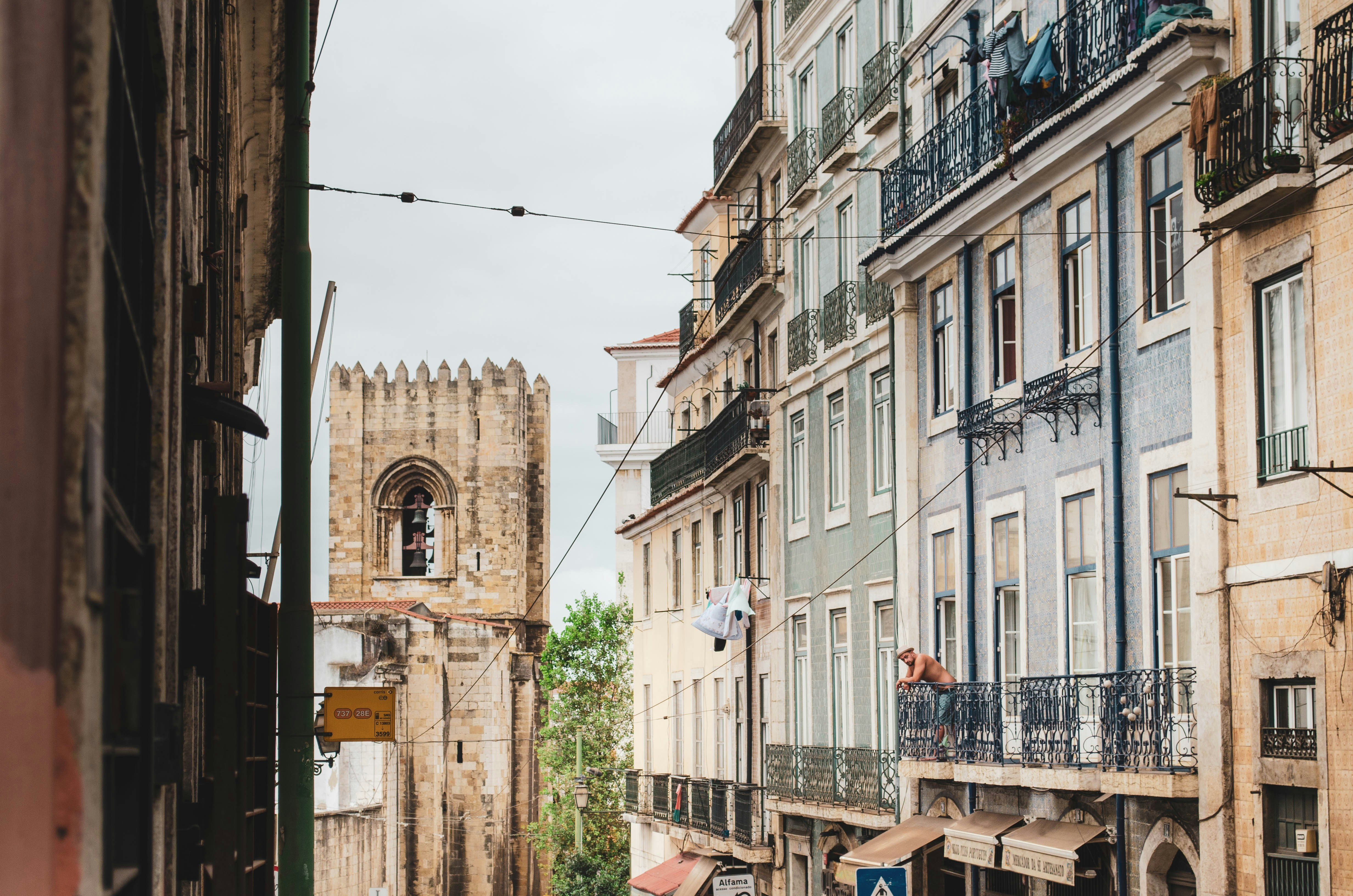 Historic buildings frame a narrow street in Lisbon, featuring a prominent bell tower and residents engaging in daily life.