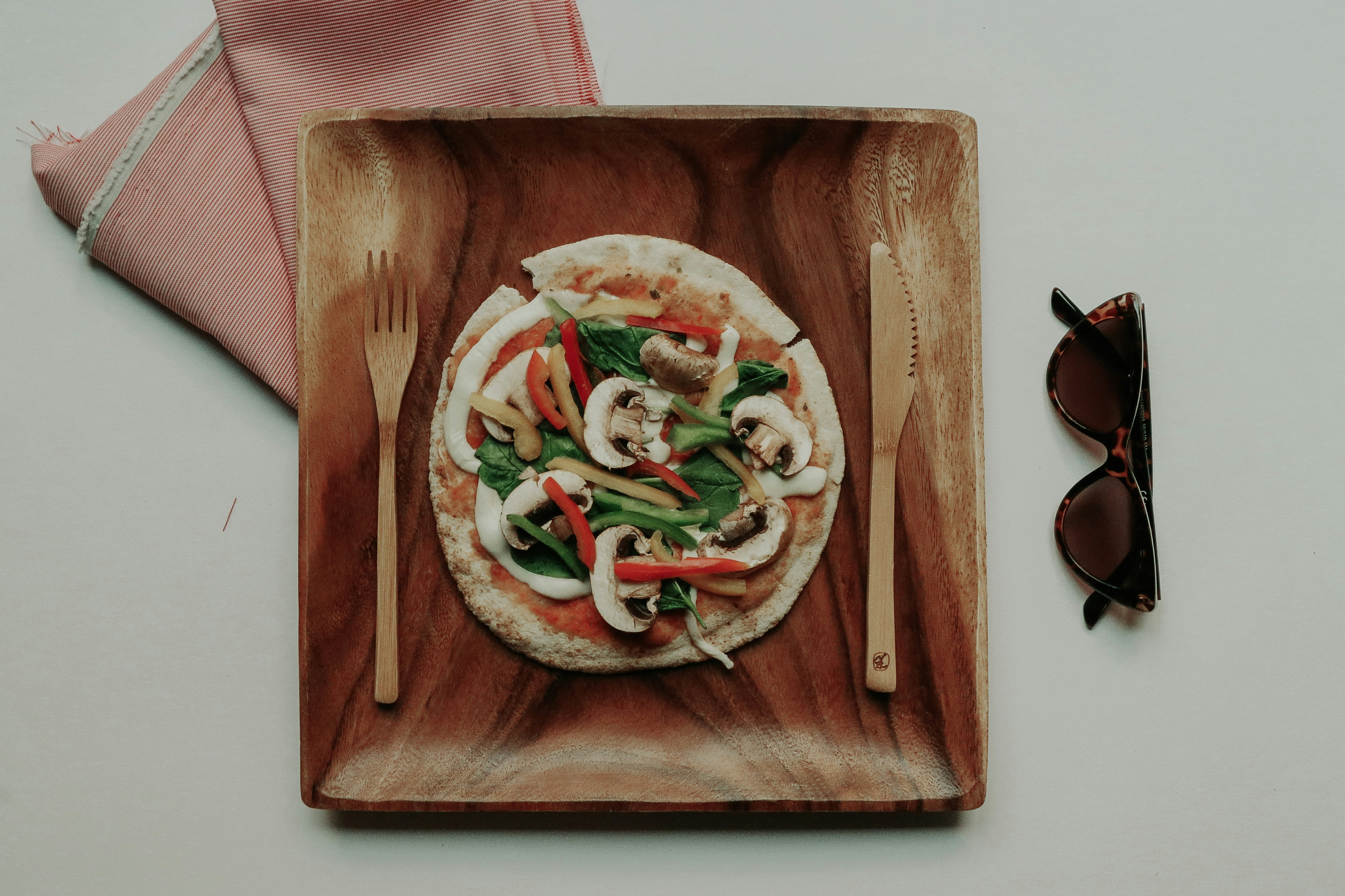 Wooden plate with vegetable pizza on a tray, framed by a pink napkin and sunglasses