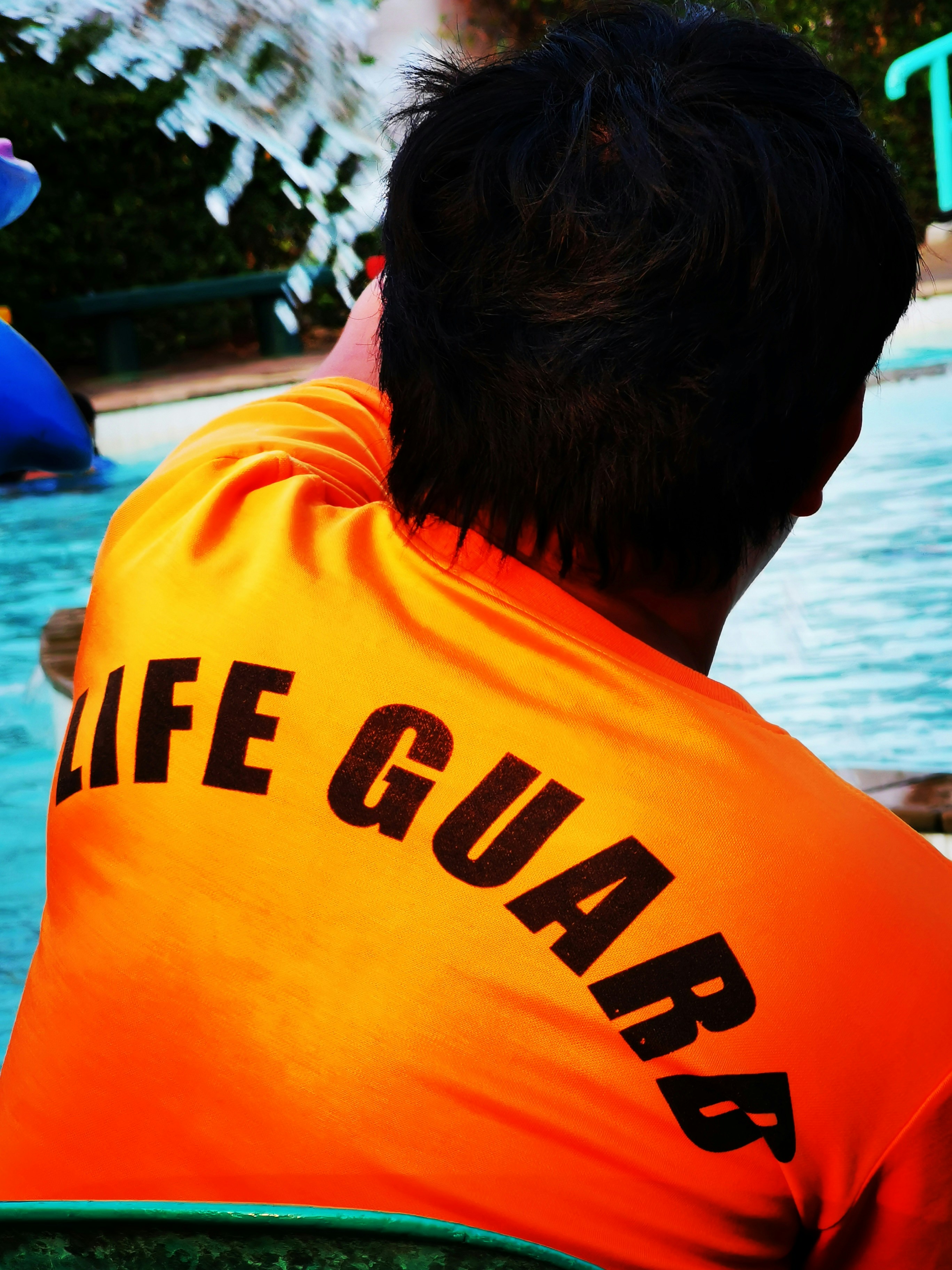 Bright orange lifeguard shirt dominates the frame as the subject faces away toward a pool, with water and poolside elements creating a vivid, candid scene.