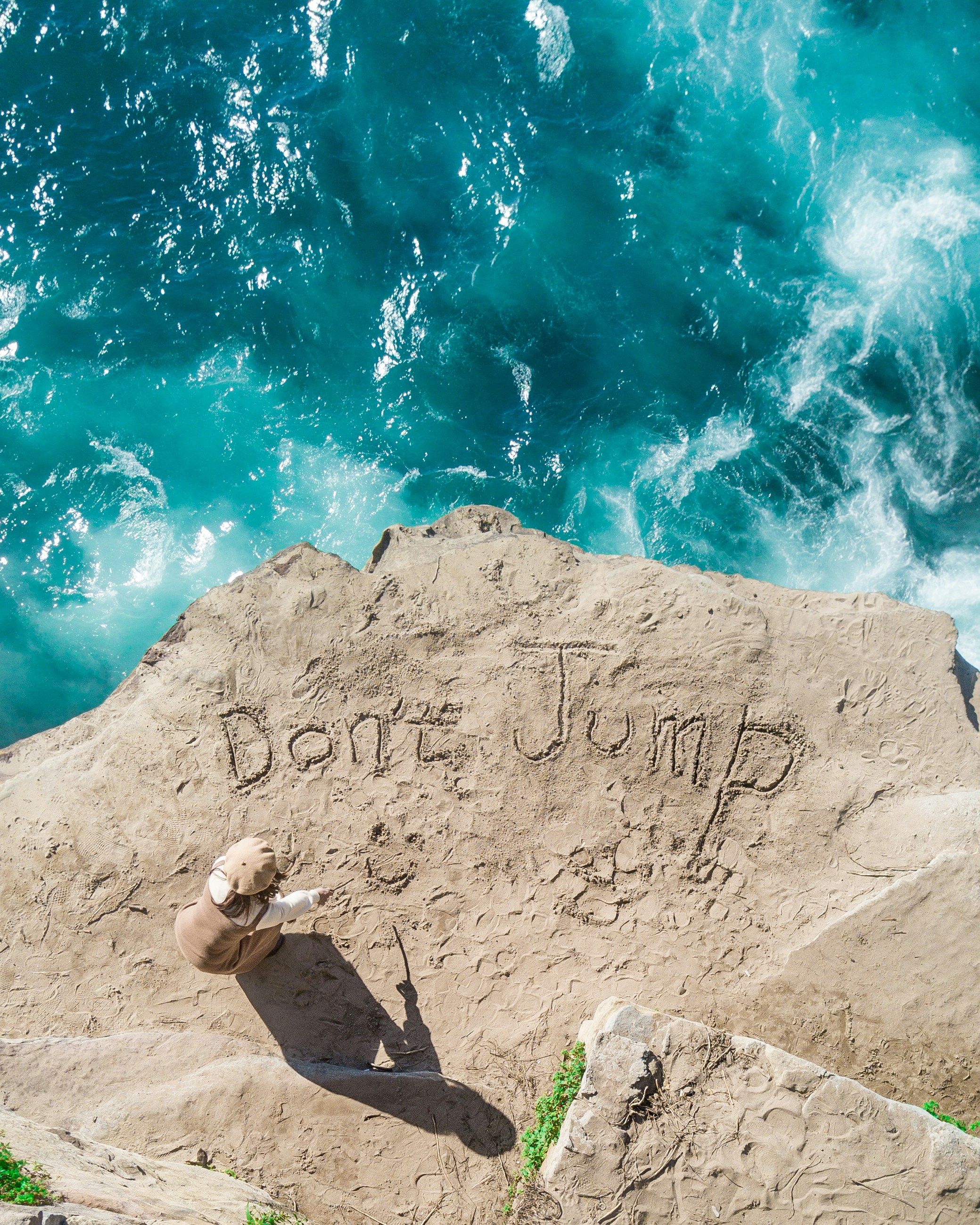 Person standing on cliff edge with 'Don't Jump' carved into the rock, overlooking vibrant blue ocean waves.