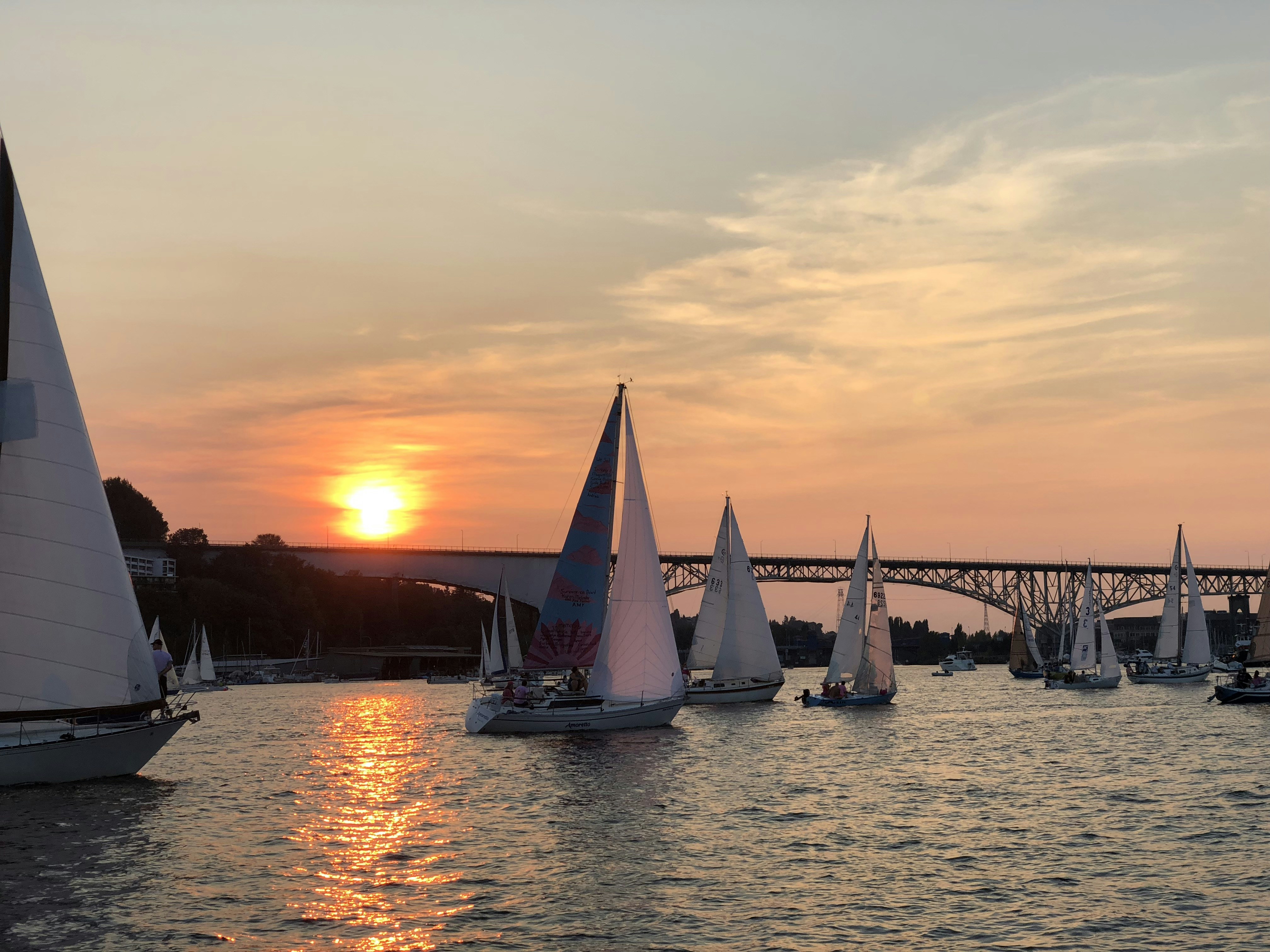 Sailboats glide under an iron bridge during a vibrant sunset.