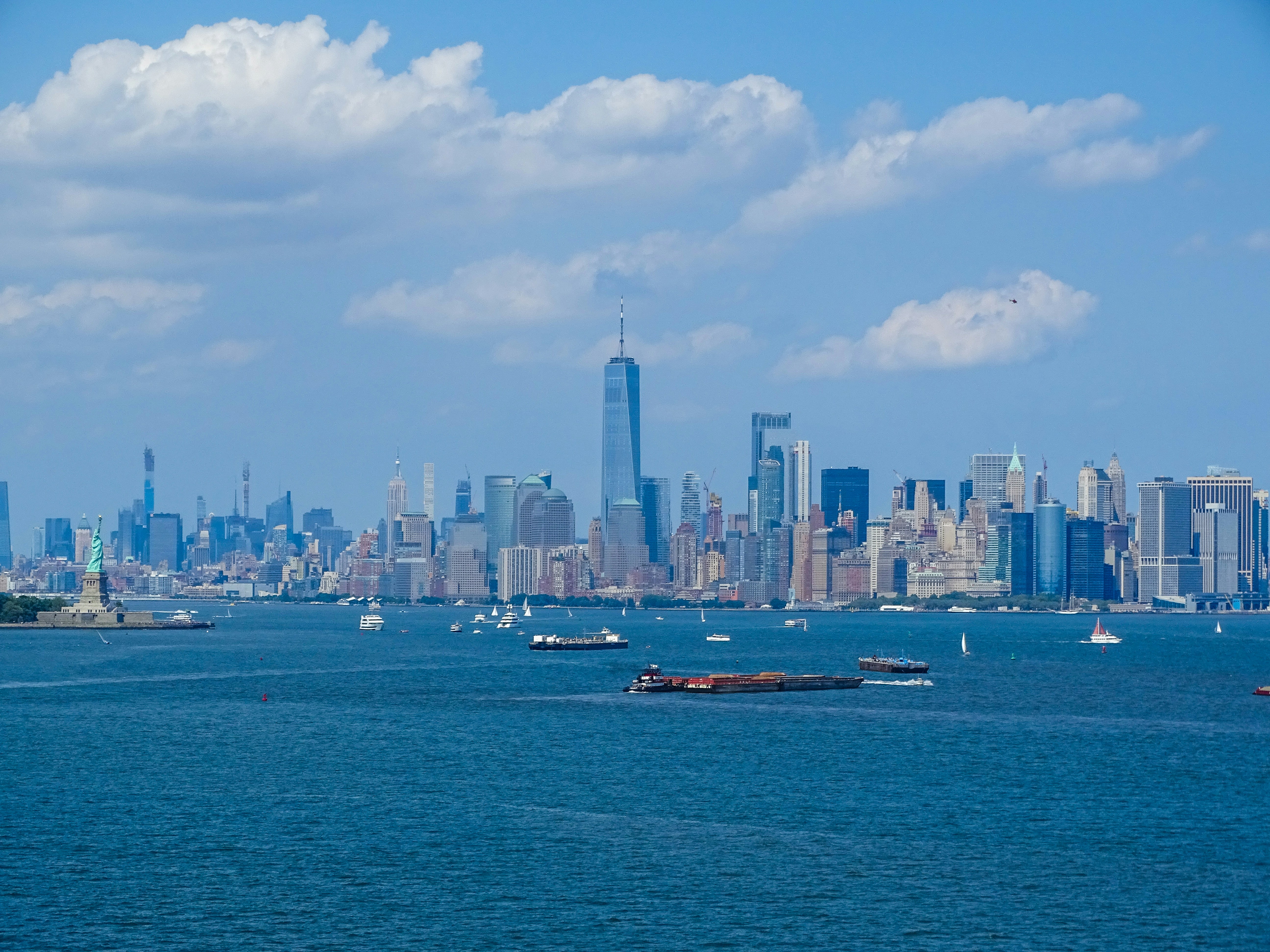 Iconic skyline of New York City featuring One World Trade Center, framed by a vibrant blue sky and bustling harbor activity.