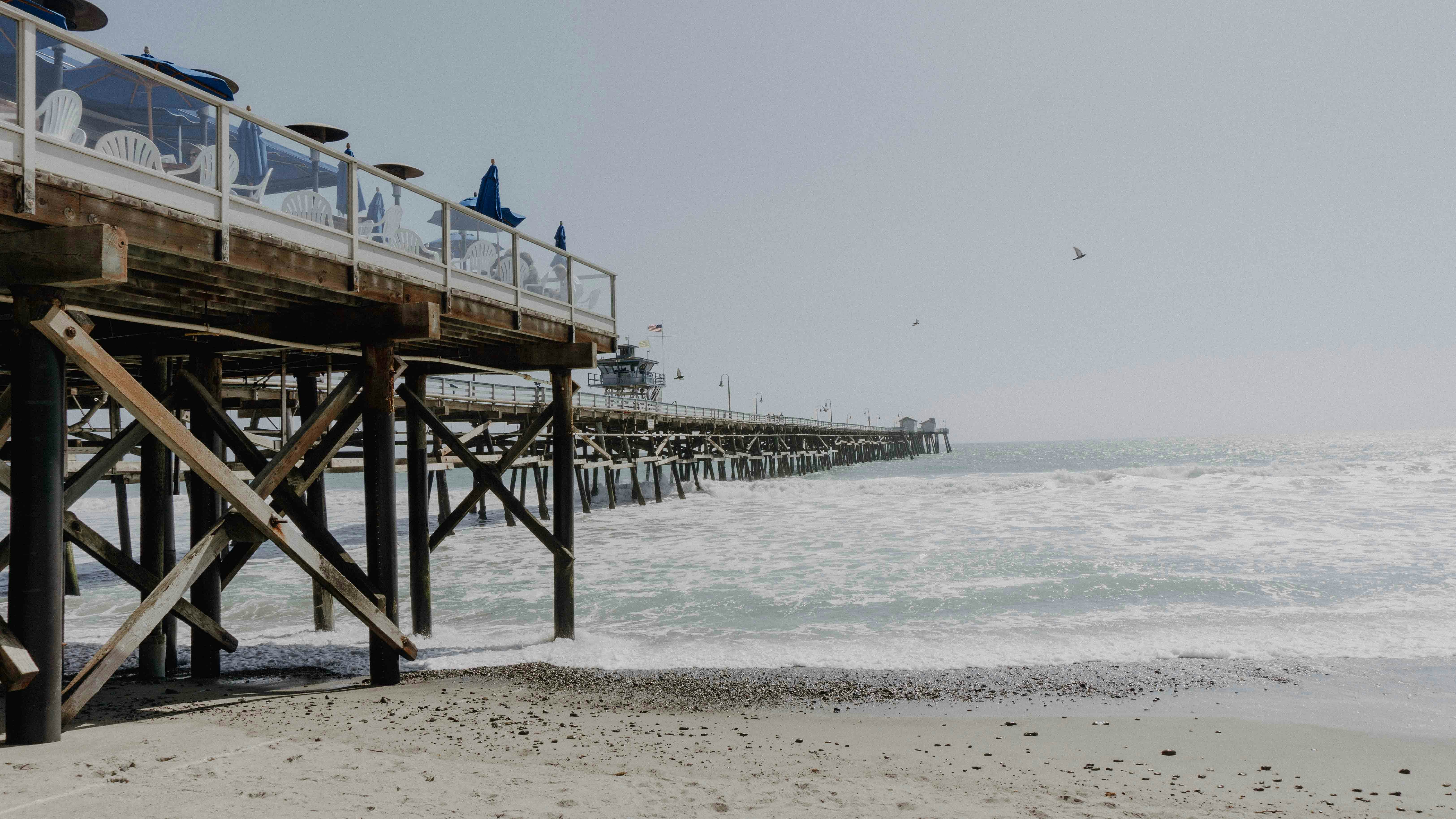 Wooden pier extending over a sunlit beach with gentle waves lapping at the shore.
