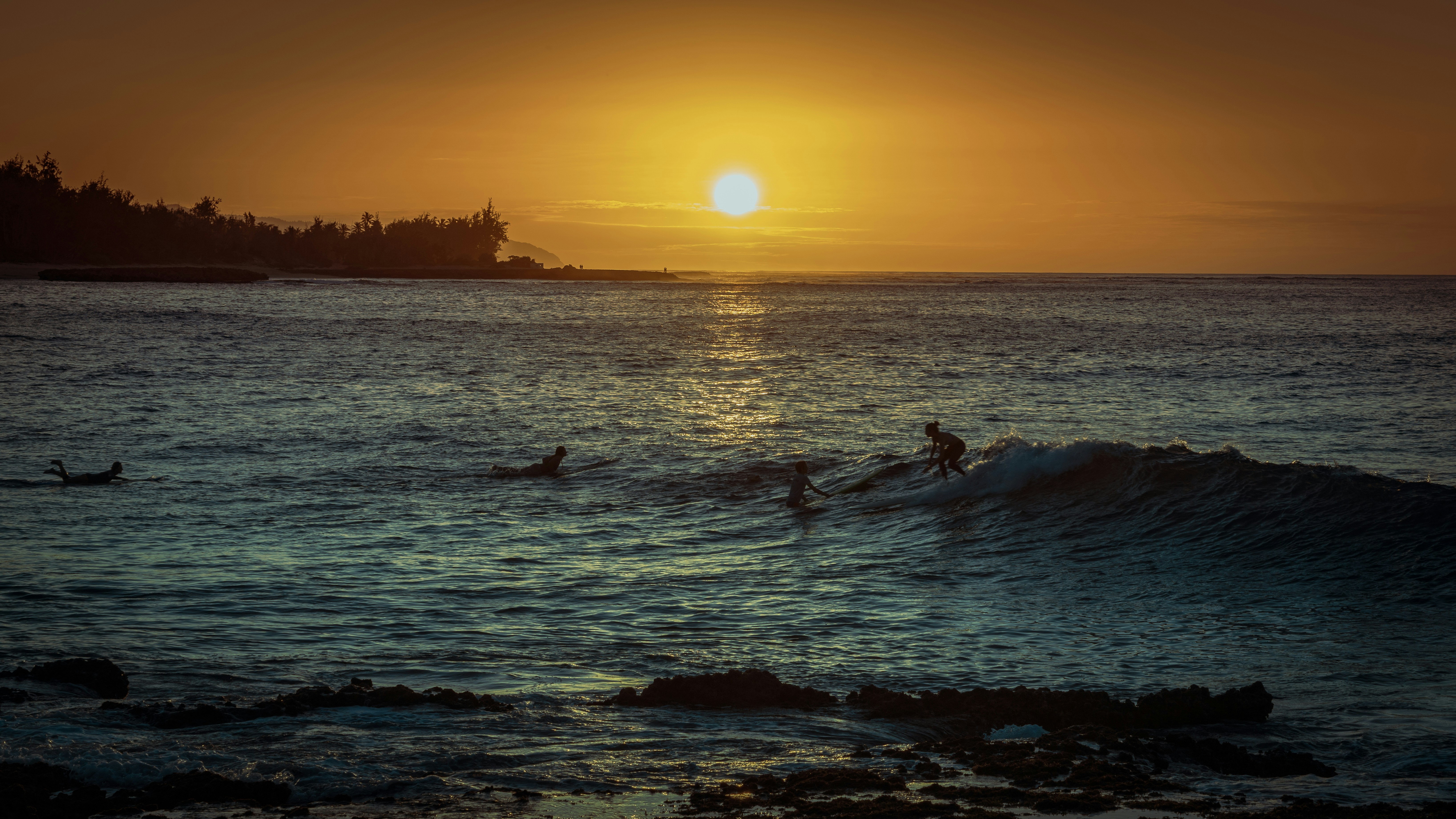 Surfers riding gentle waves as the sun sets over the horizon, casting a warm glow on the water.