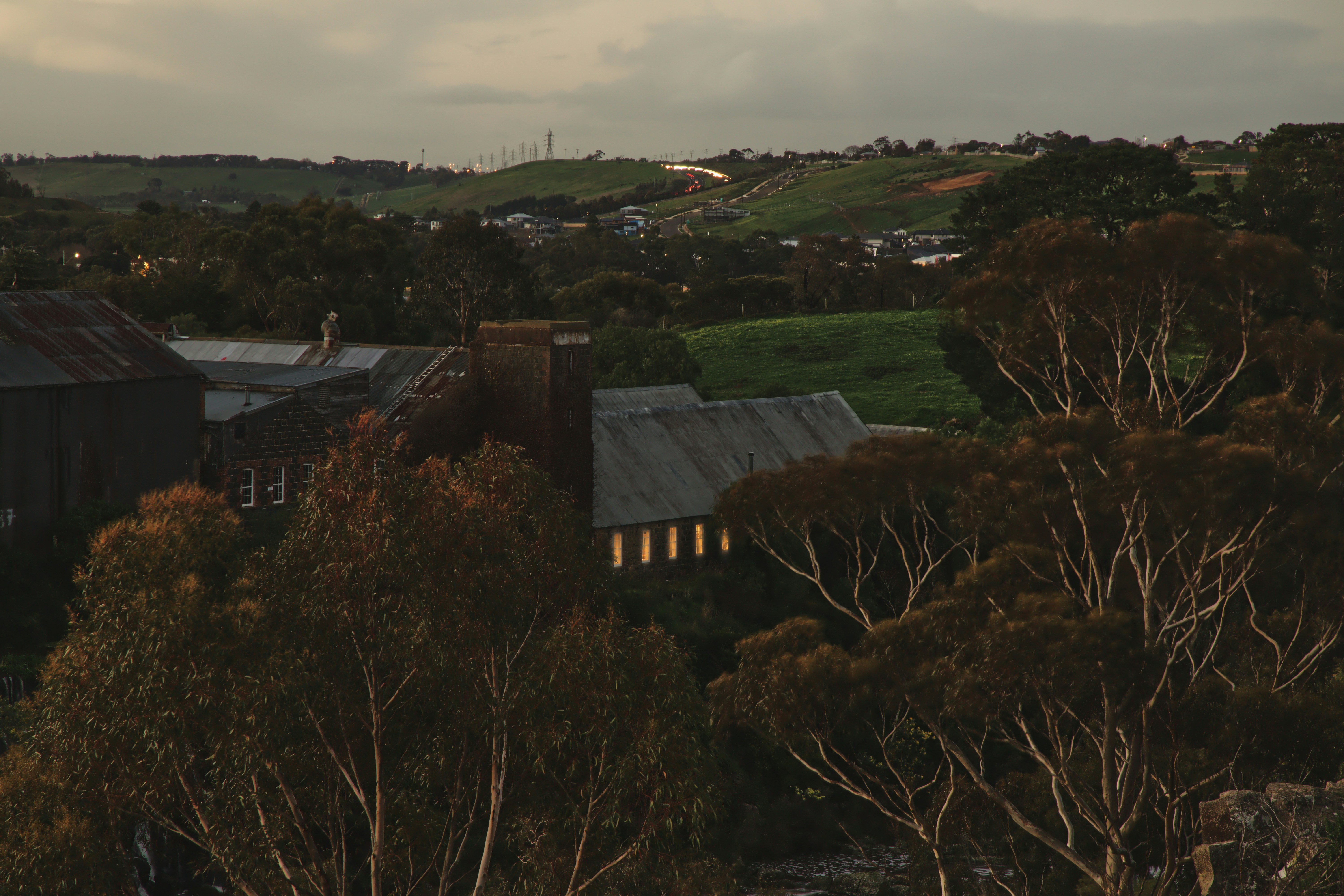 Rustic buildings nestled among trees with a distant view of rolling hills under a cloudy sky. Warm lights glow from the windows, hinting at life within.