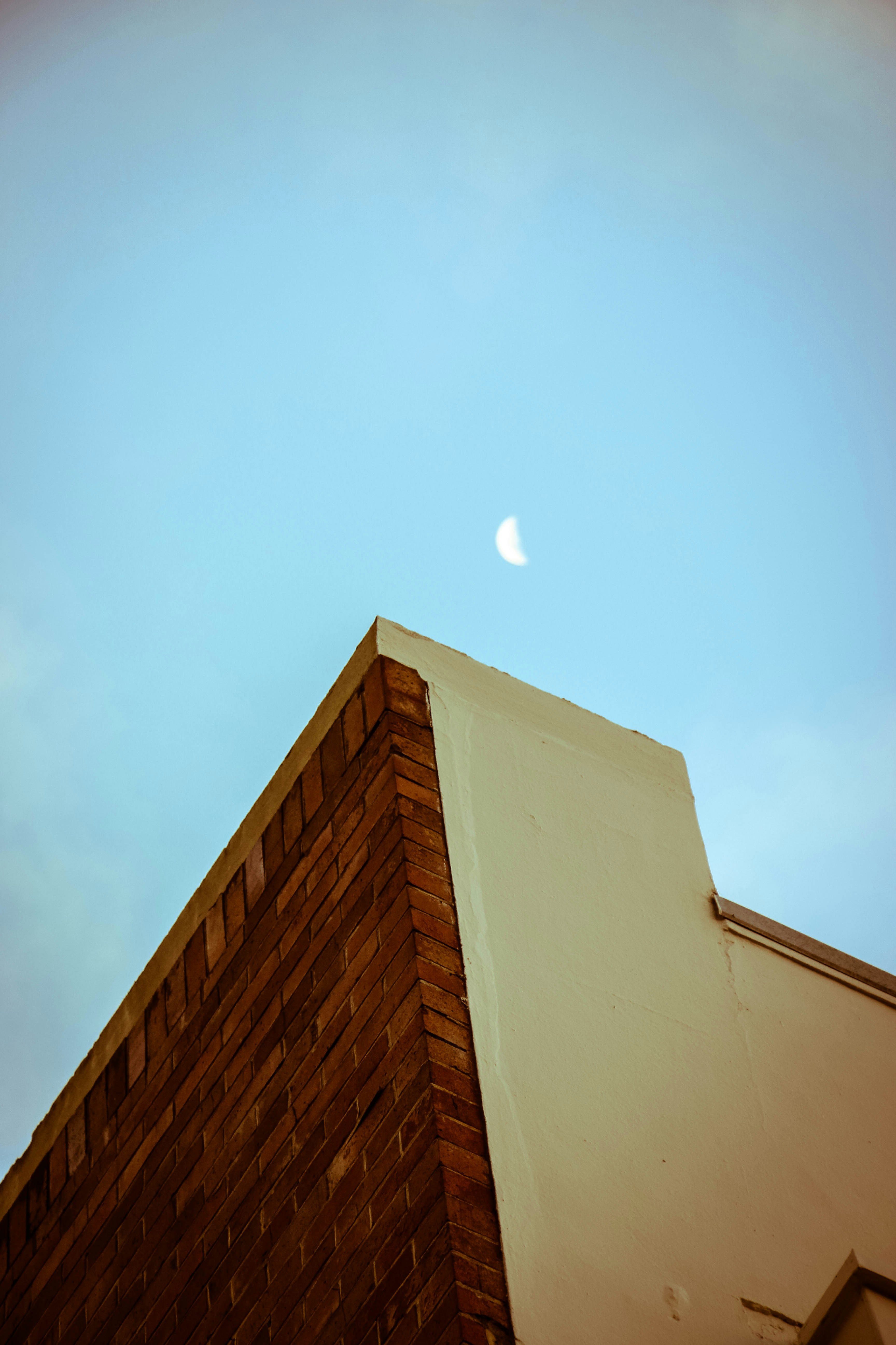 A crescent moon peeks over the edge of a brick building against a soft blue sky.
