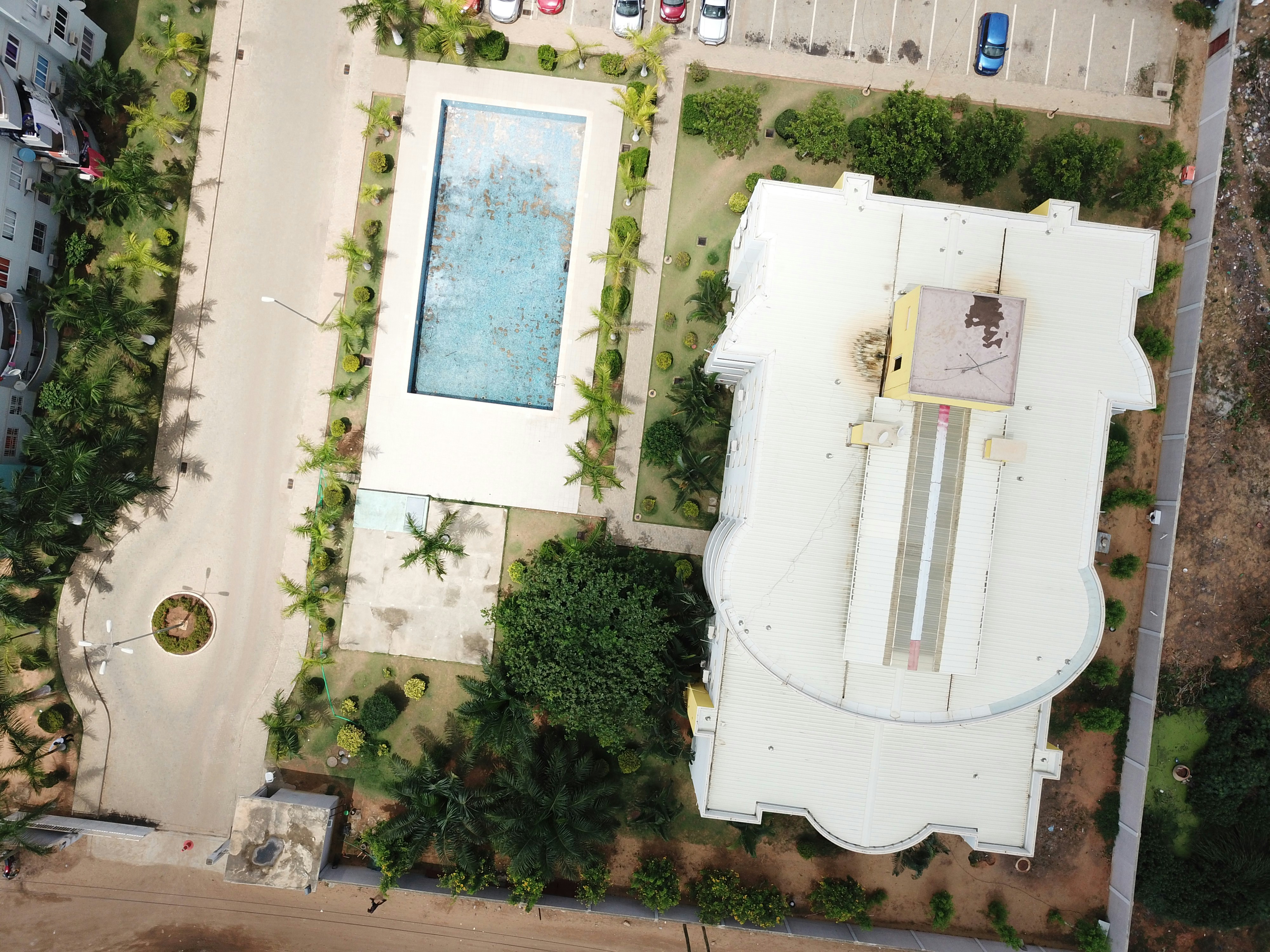 Aerial view of a white building adjacent to a rectangular swimming pool surrounded by greenery.
