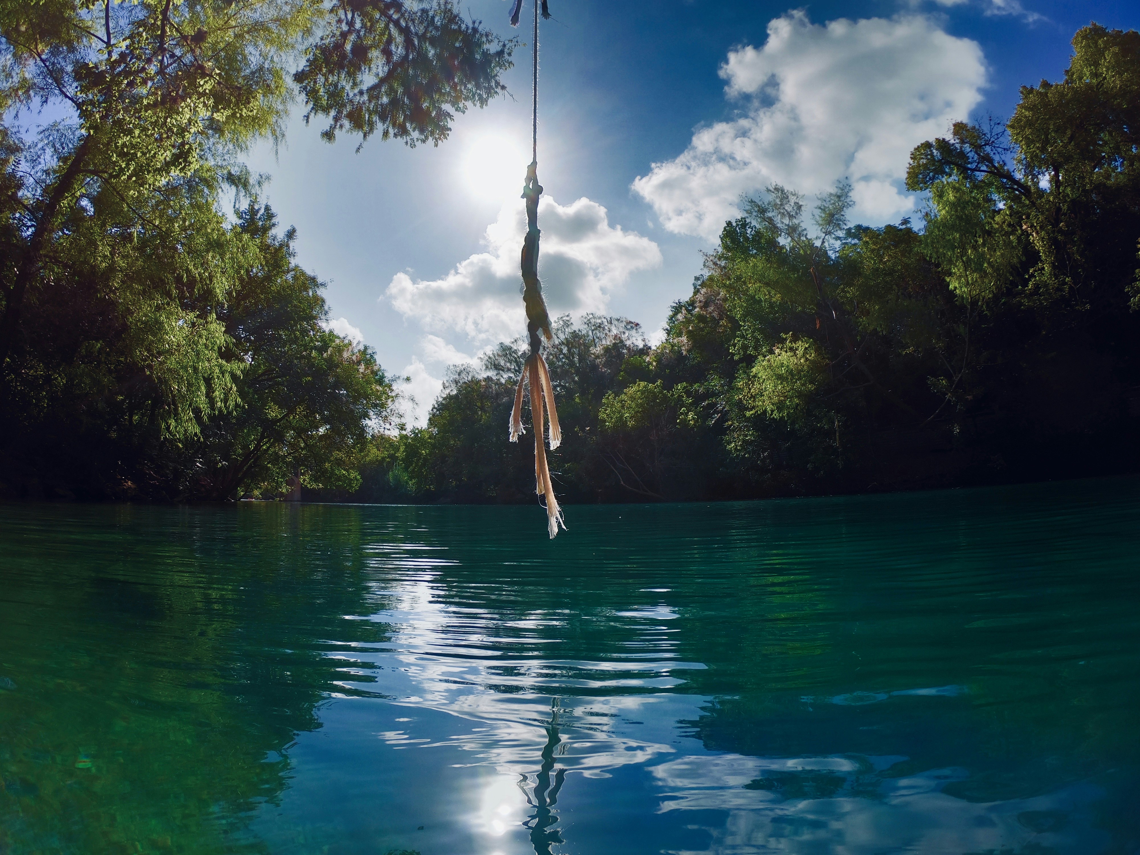 Person swinging over a tranquil river surrounded by lush trees and a bright sunlit sky.