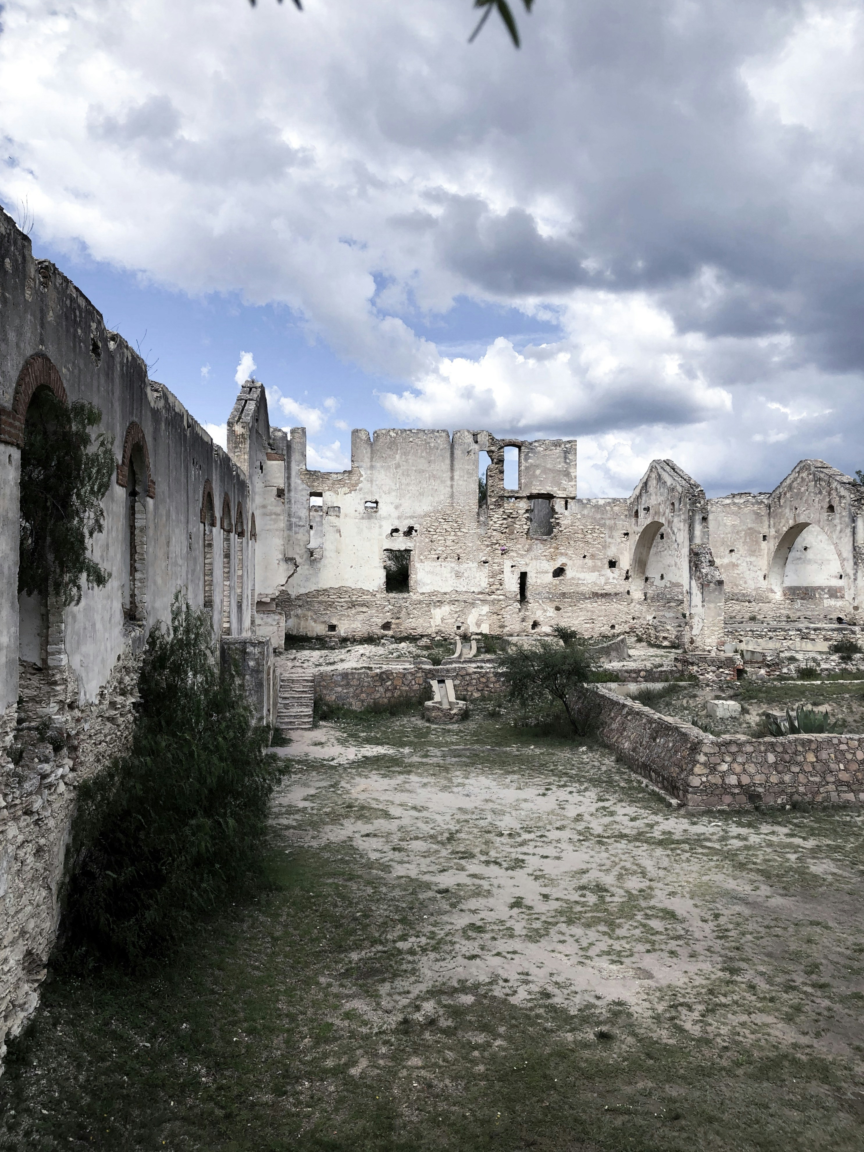 Ancient ruins of a stone structure surrounded by overgrown grass and trees under a dramatic sky.