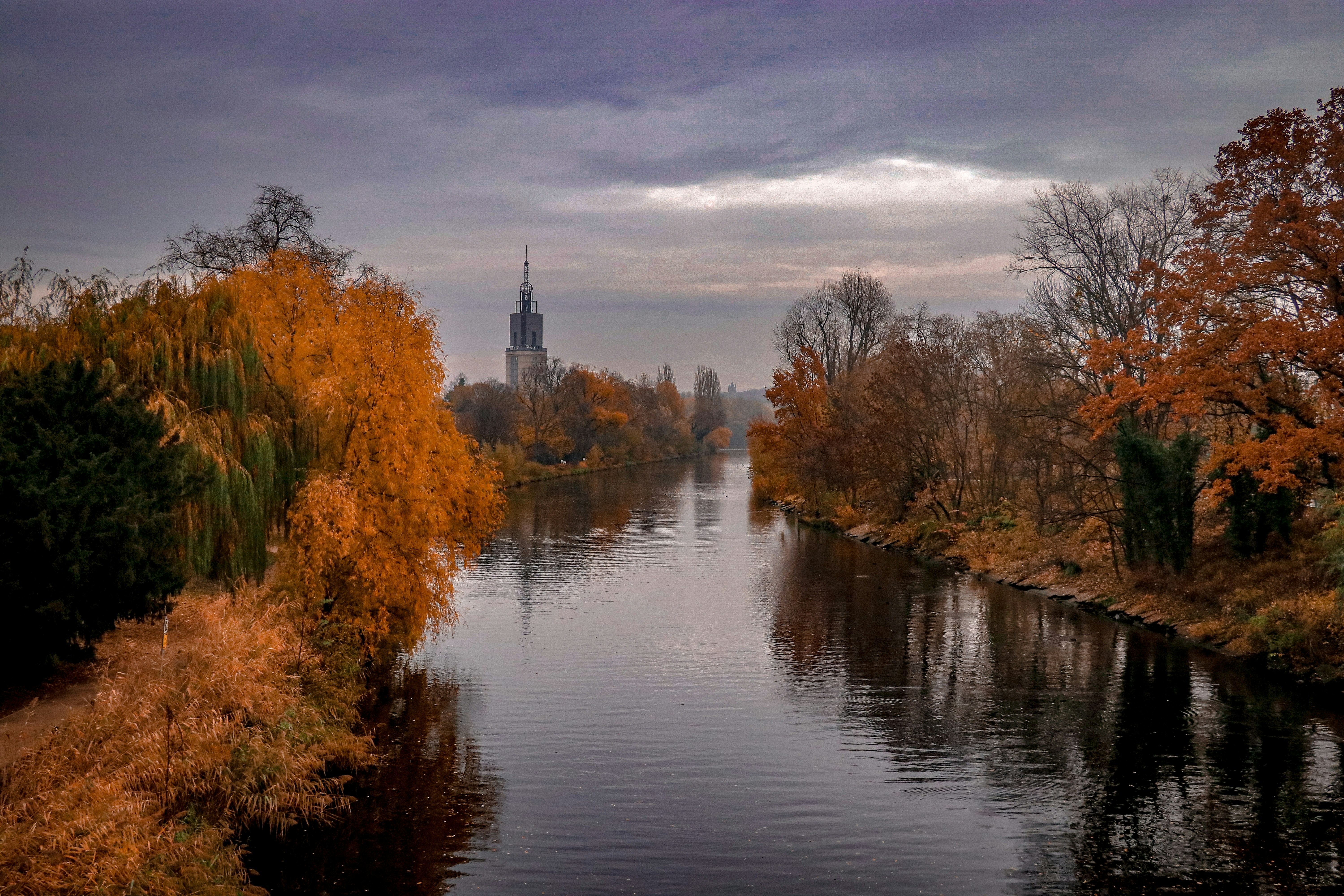 Serene river flanked by vibrant autumn trees, with a distant church spire under a cloudy sky.