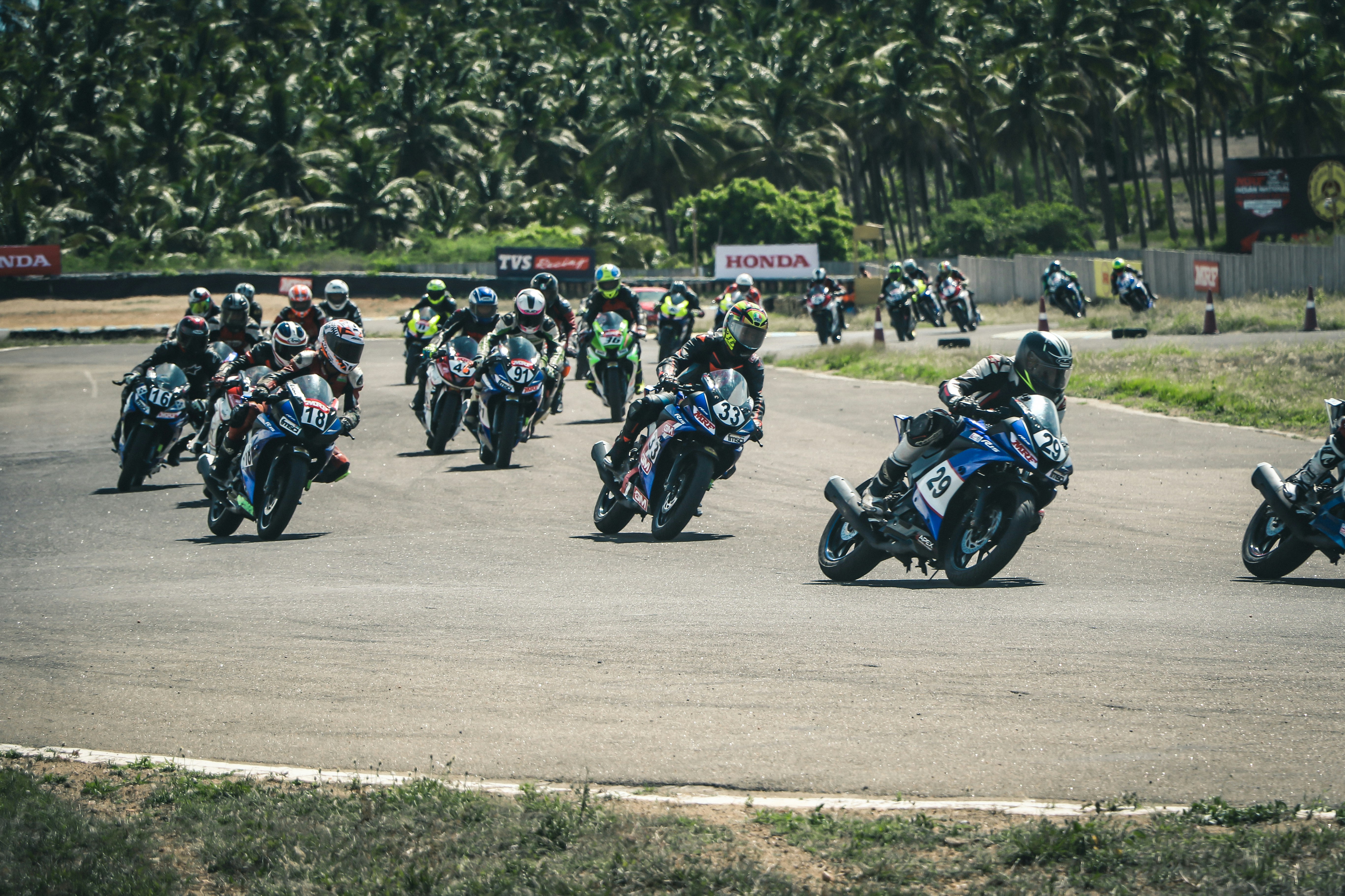 Motorcyclists lean into a sharp curve on a race track surrounded by lush greenery under a cloudy sky.