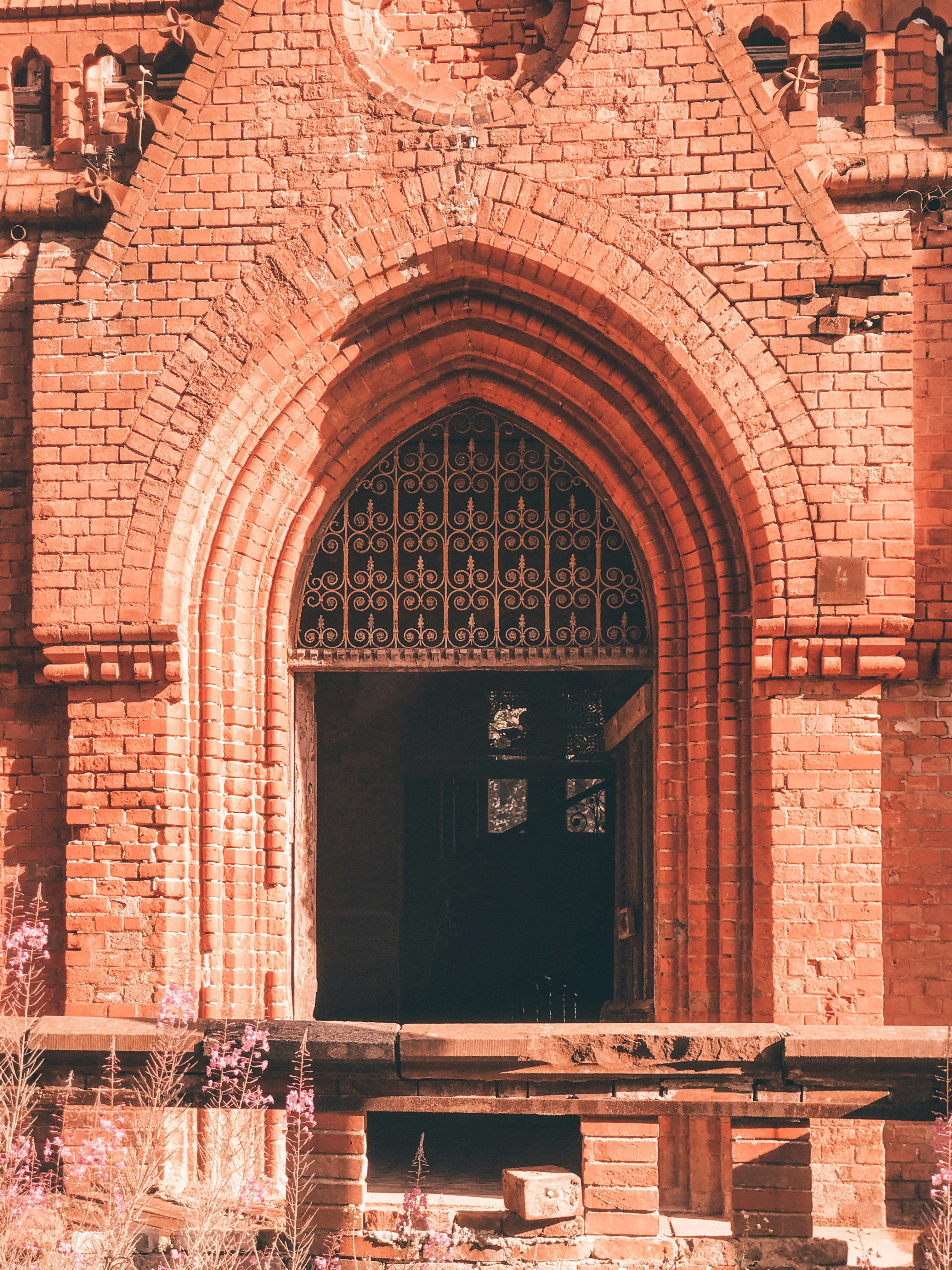 Intricate brick archway with ornate ironwork leading to a darkened interior, framed by wildflowers in the foreground.