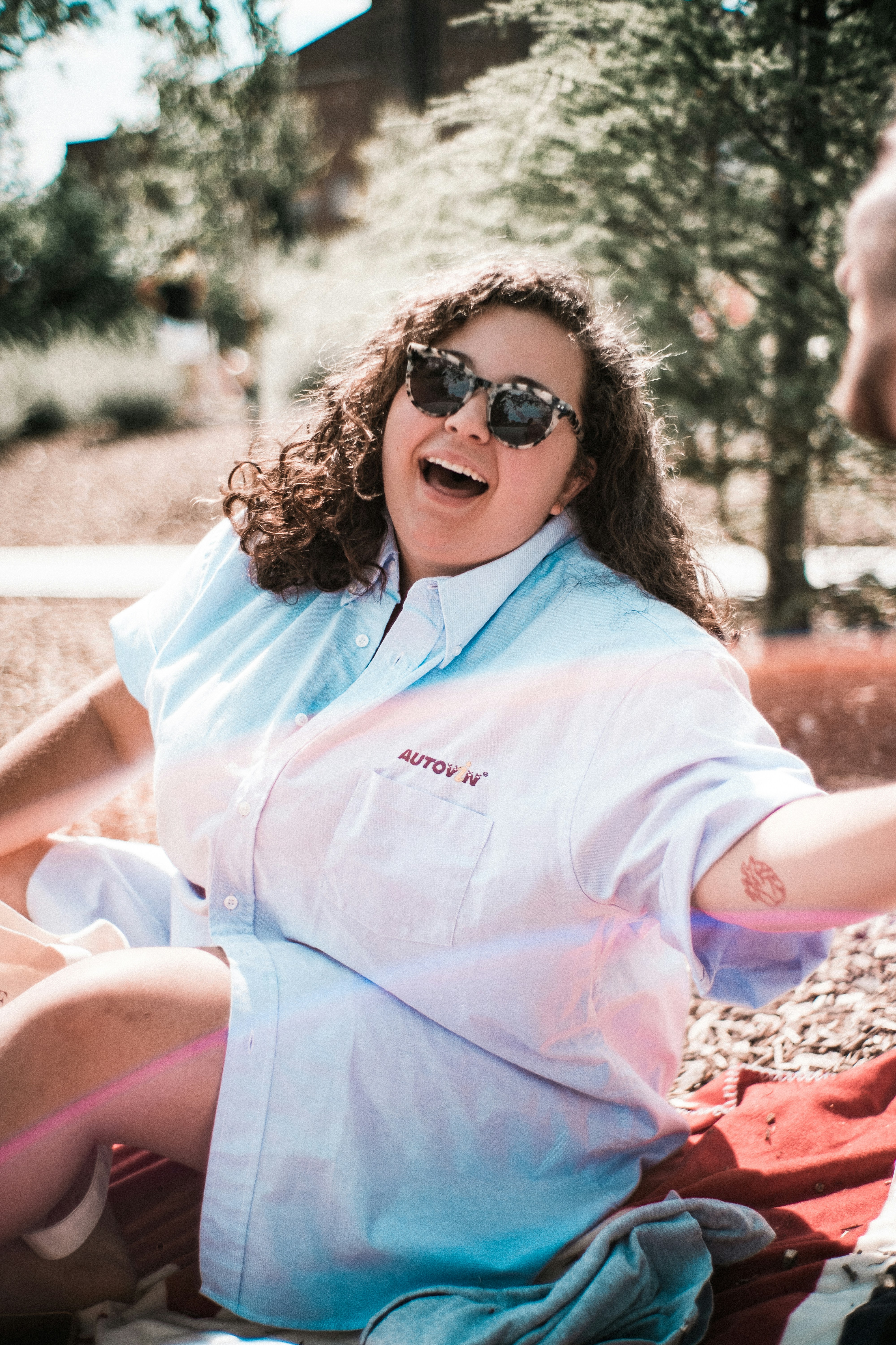 Woman in oversized shirt laughing while sitting outdoors, surrounded by greenery and sunlight.