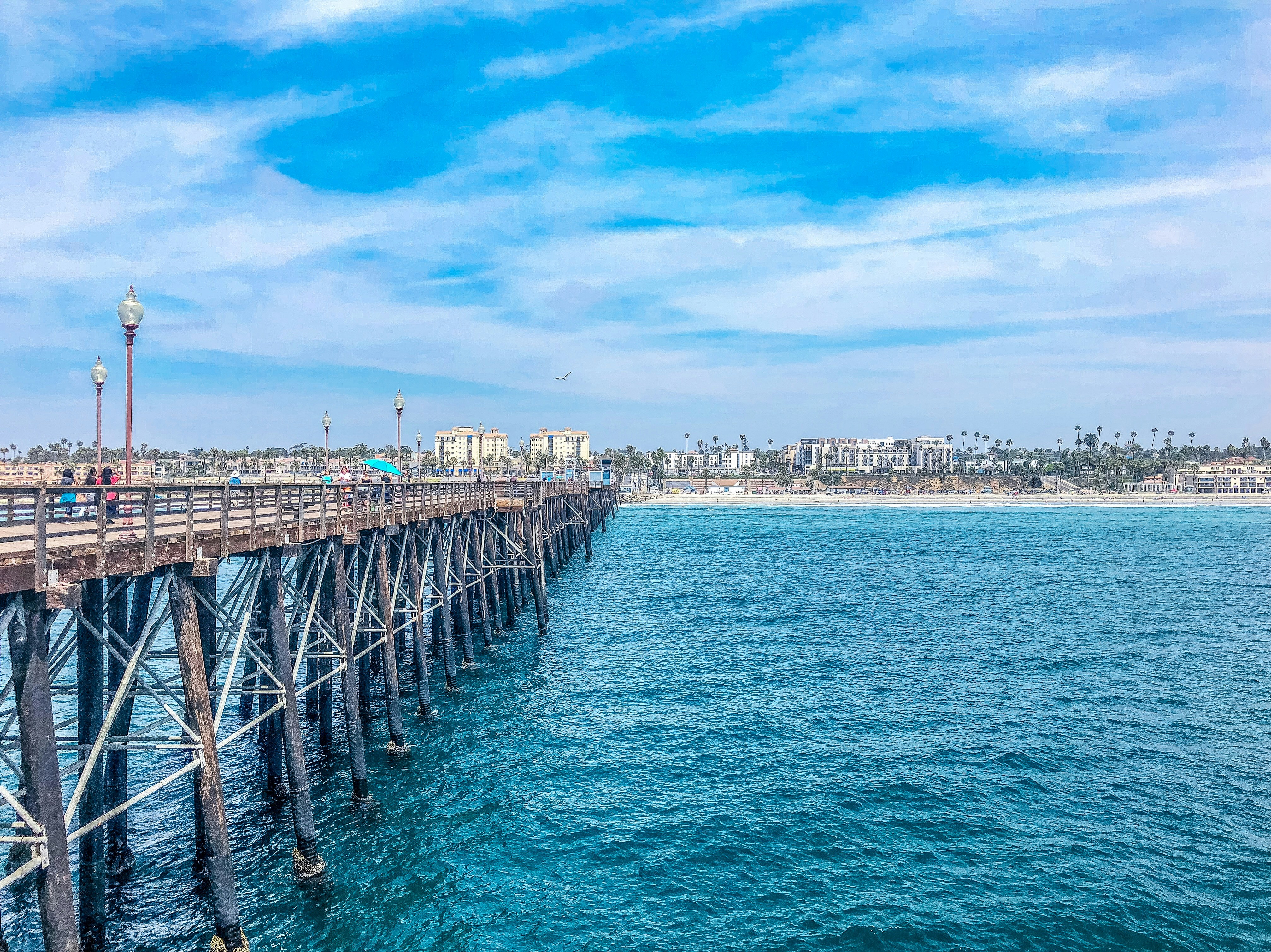 Wooden pier stretching into a vibrant blue ocean with a distant shoreline under a sky filled with wispy clouds.
