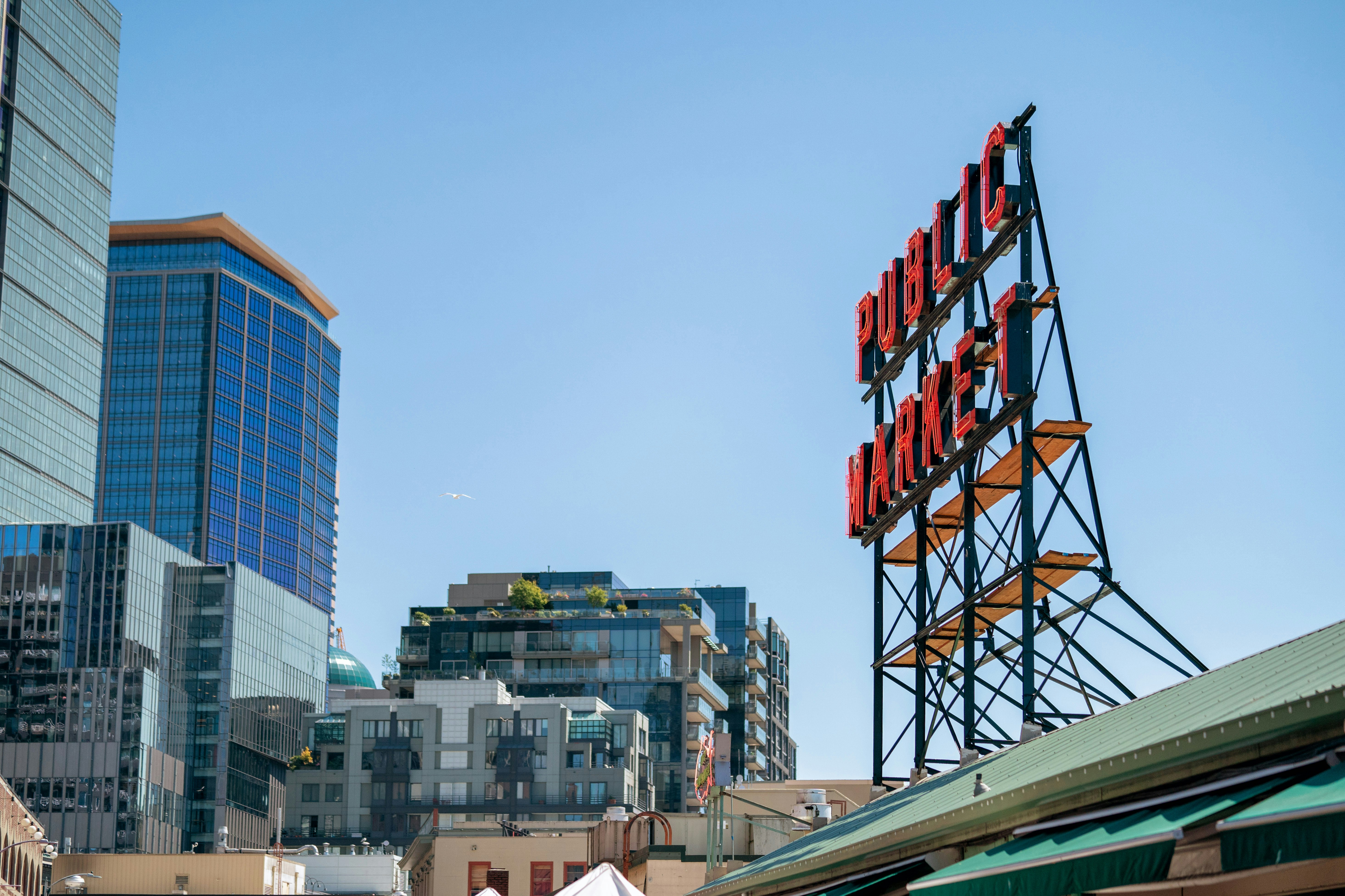 Iconic 'PUBLIC MARKET' sign towers above a vibrant cityscape, showcasing a blend of historic and modern architecture. Bright blue sky enhances the urban atmosphere.
