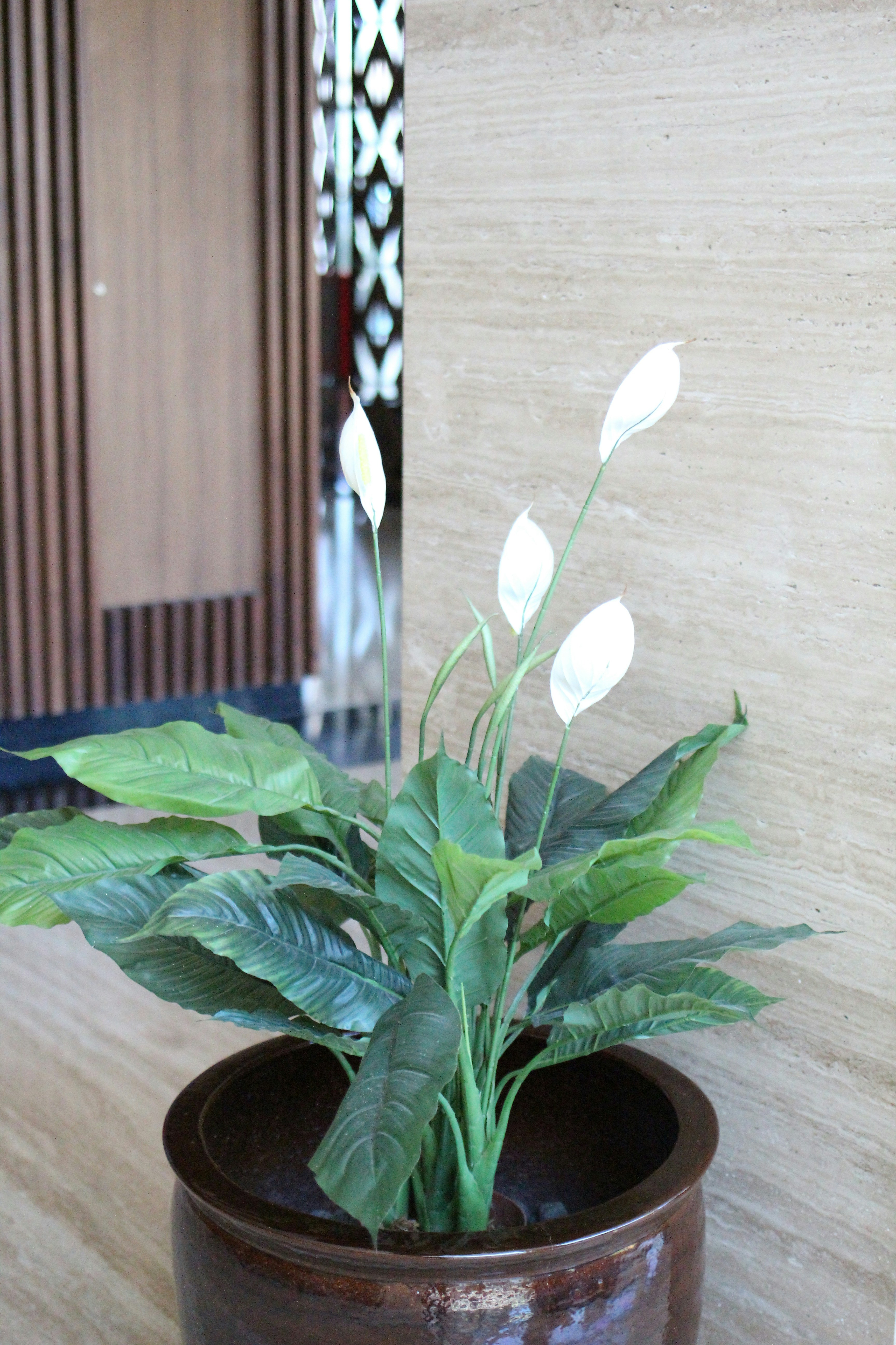 Artificial peace lily plant arranged in a brown pot, set against a textured stone wall, enhancing indoor tranquility.