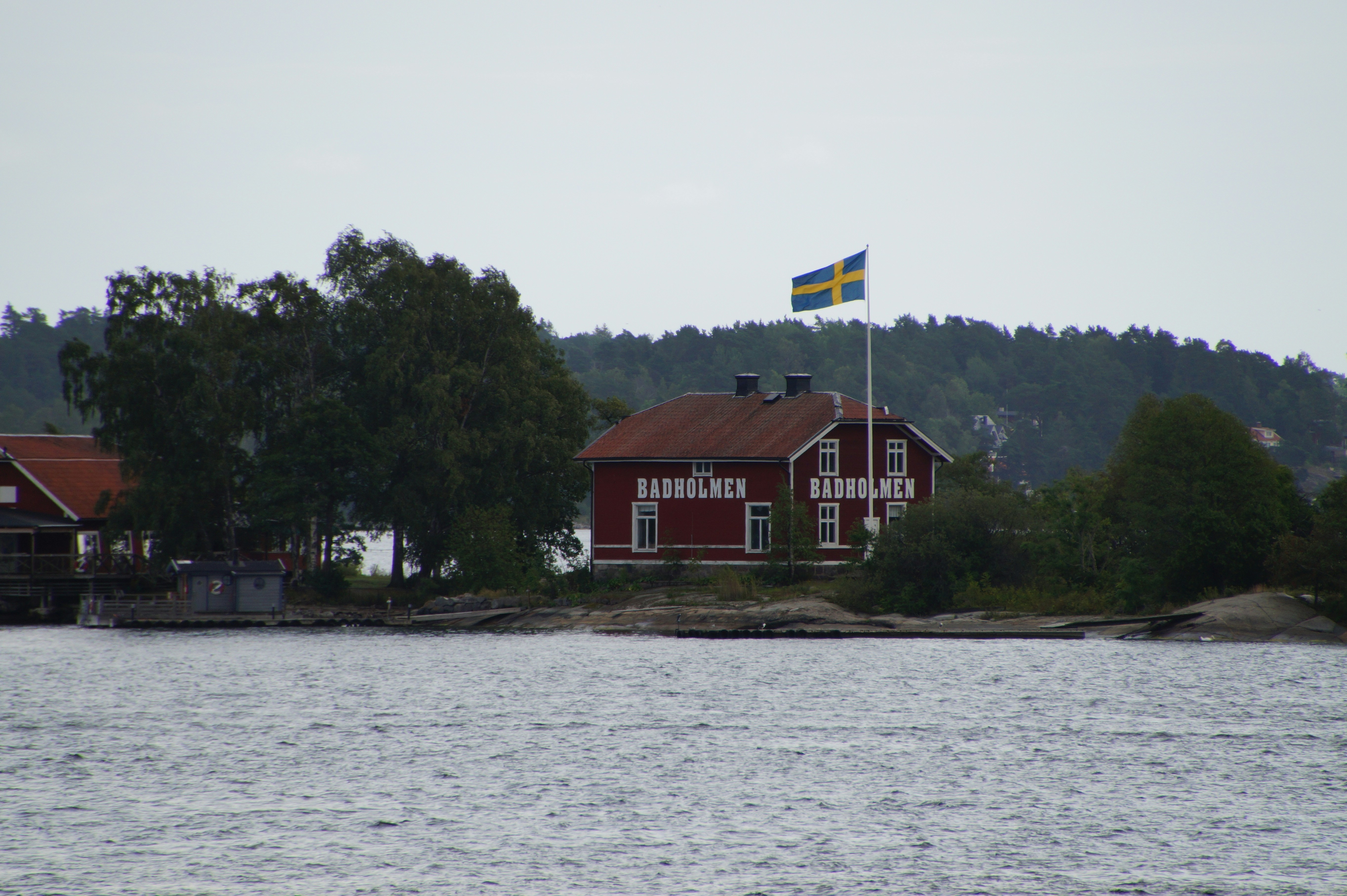 Historic red wooden building with a Swedish flag near a calm lake, surrounded by trees and distant hills.