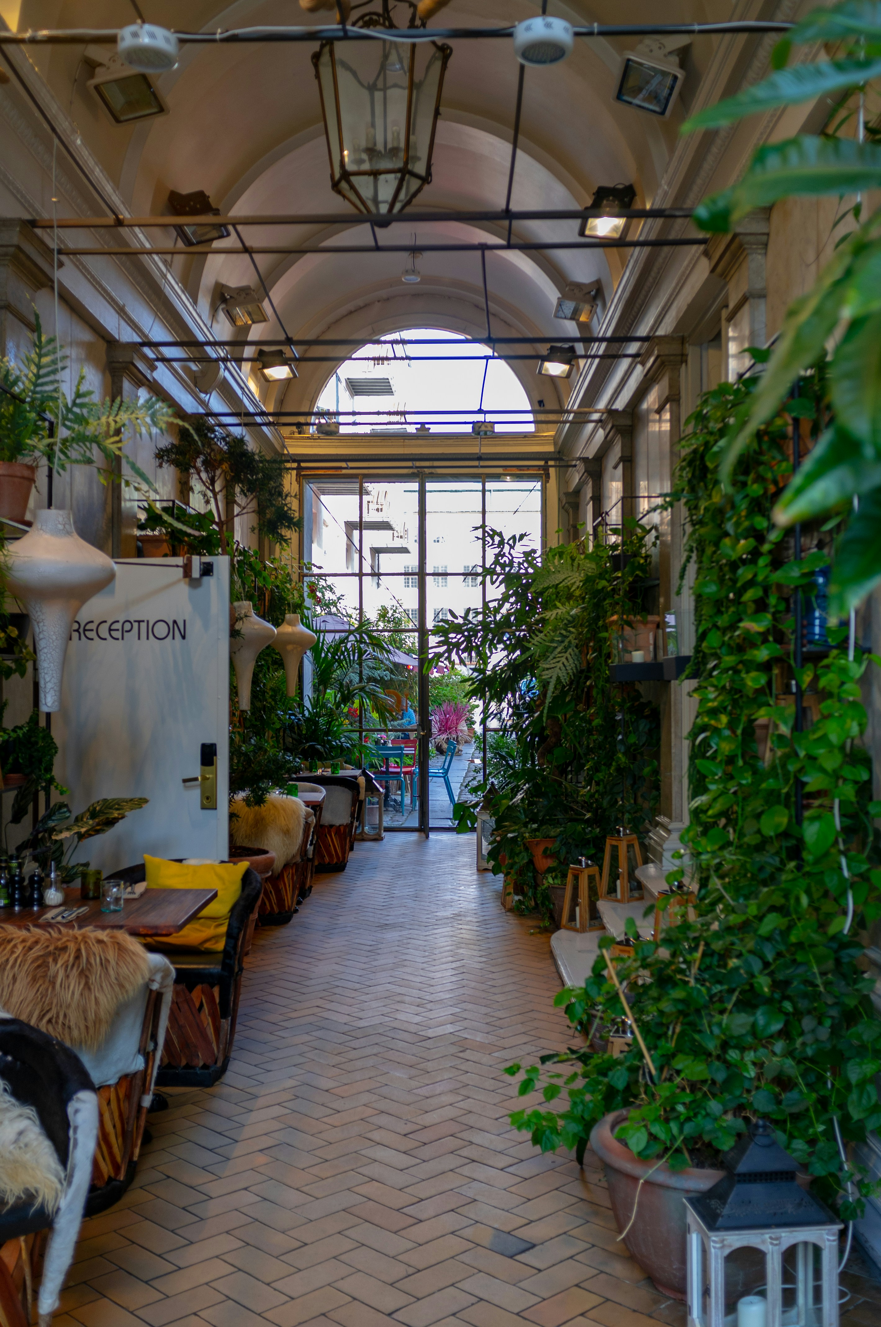 Lush indoor garden corridor featuring hanging plants and cozy seating, leading to a bright reception area. The inviting atmosphere combines nature with modern design.