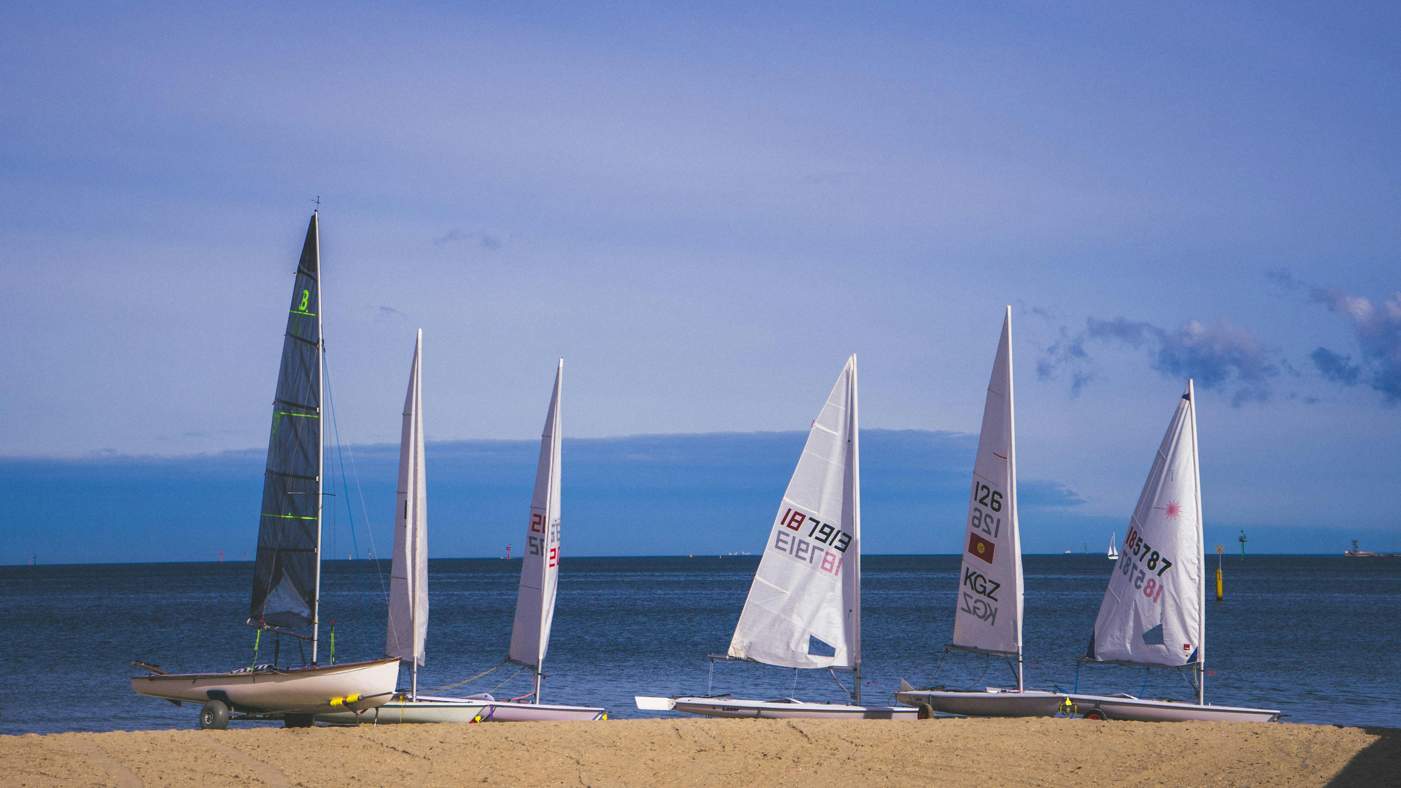A row of sailboats with colorful sails resting on a sandy beach, set against a serene blue ocean backdrop.