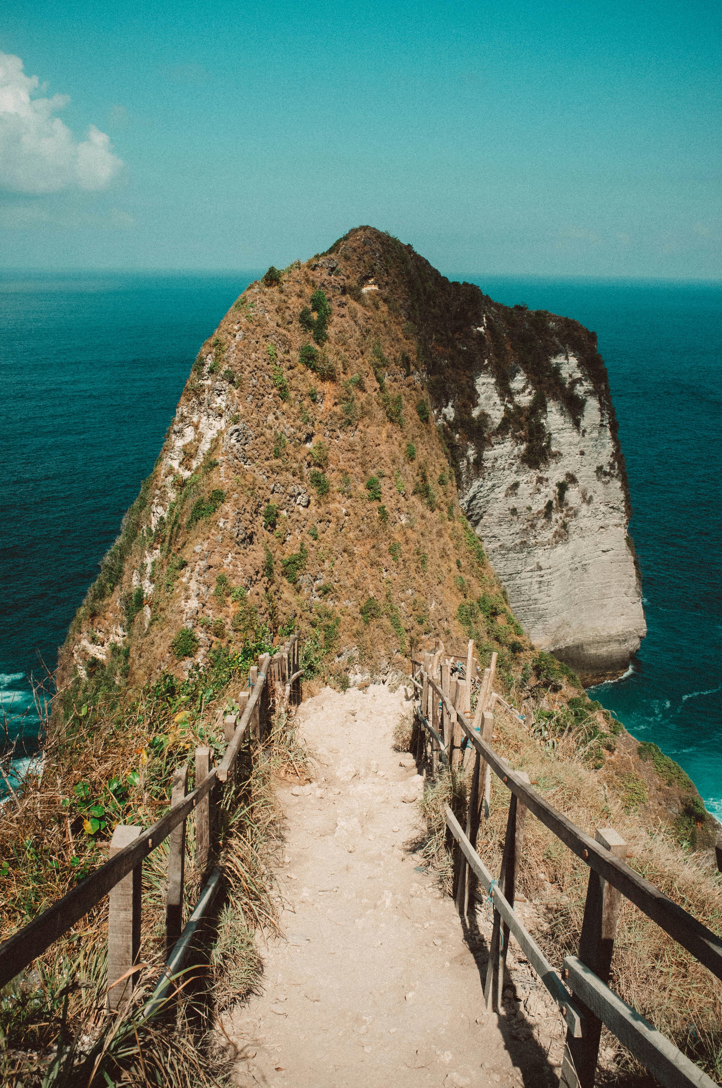 Narrow dirt path with wooden railings leading to a steep cliff surrounded by blue ocean.