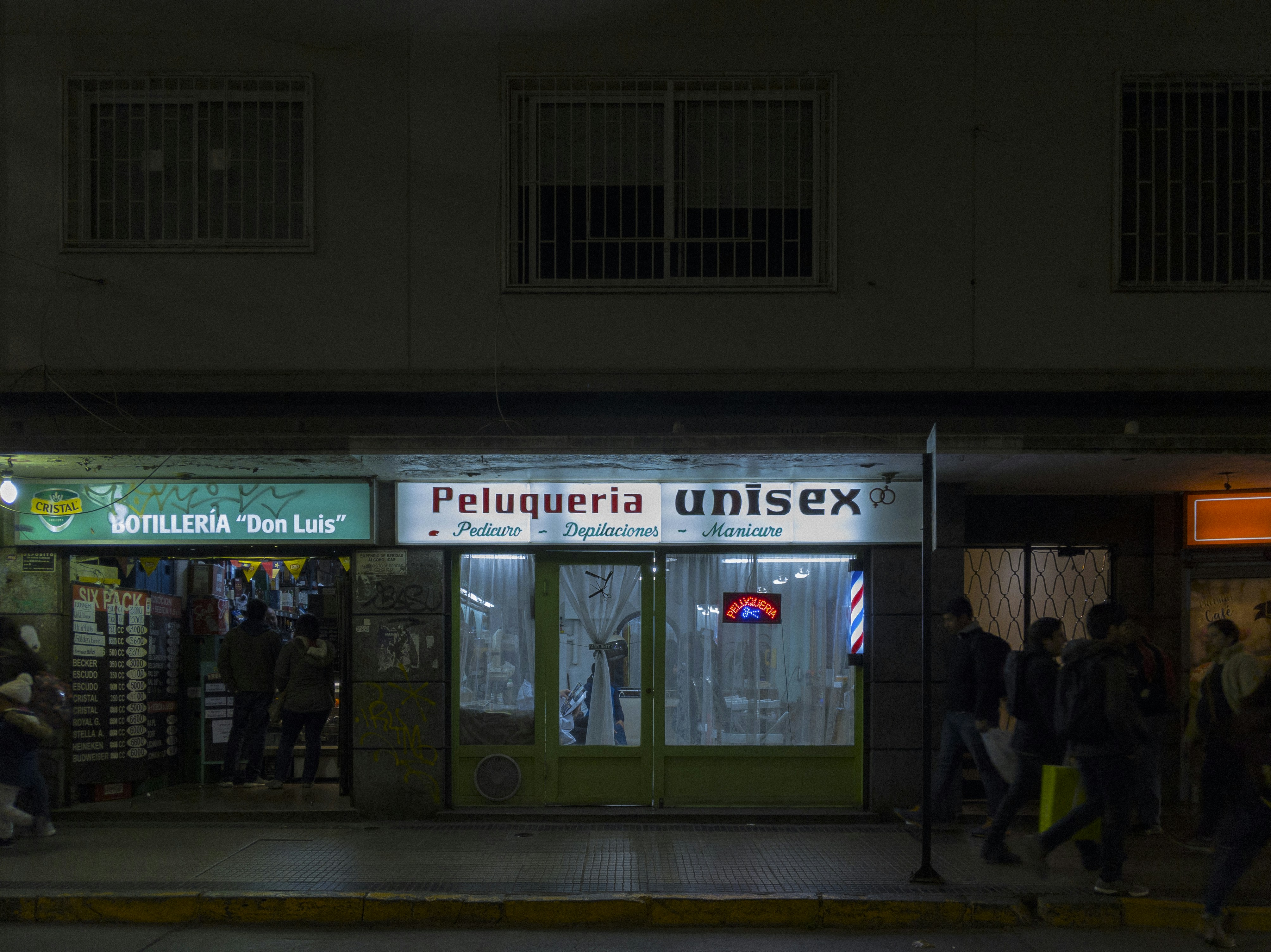Dimly lit storefront of a unisex salon with glowing neon sign, flanked by a nearby shop and passersby.