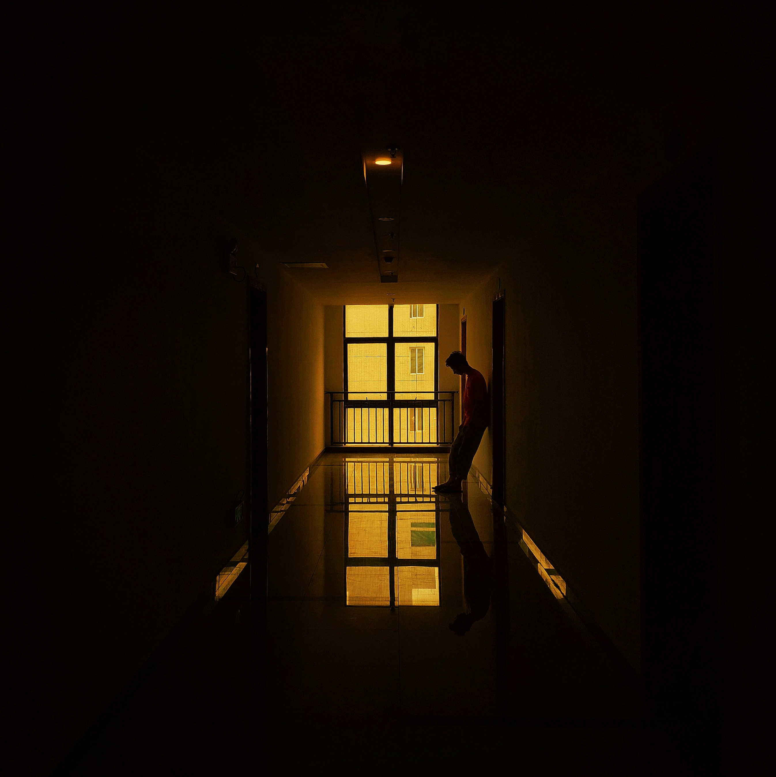 Person leaning in silhouette against a window at the end of a dimly lit hallway with golden reflections.