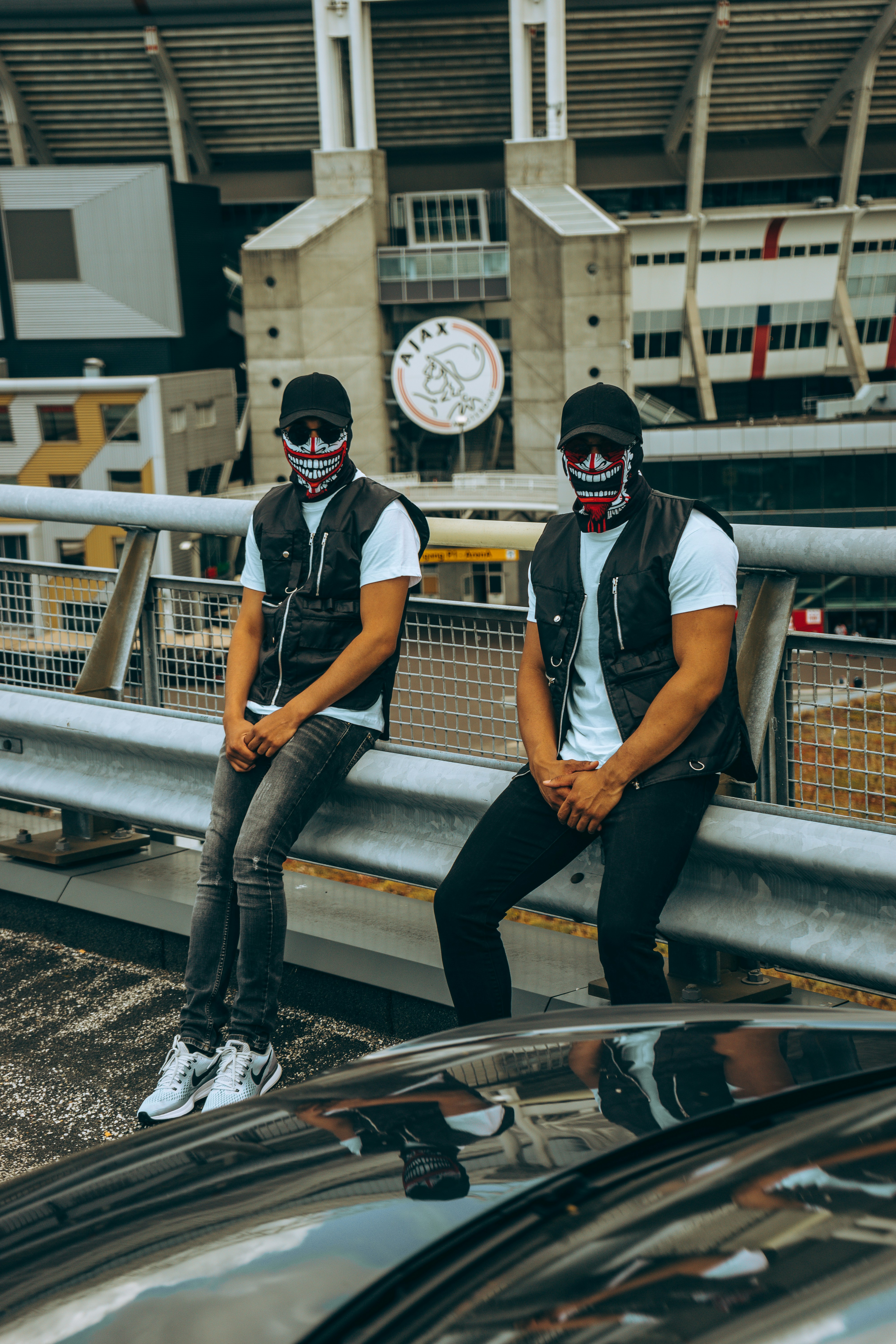 Twin masked riders on a city overlook, mirrored in a glossy car hood