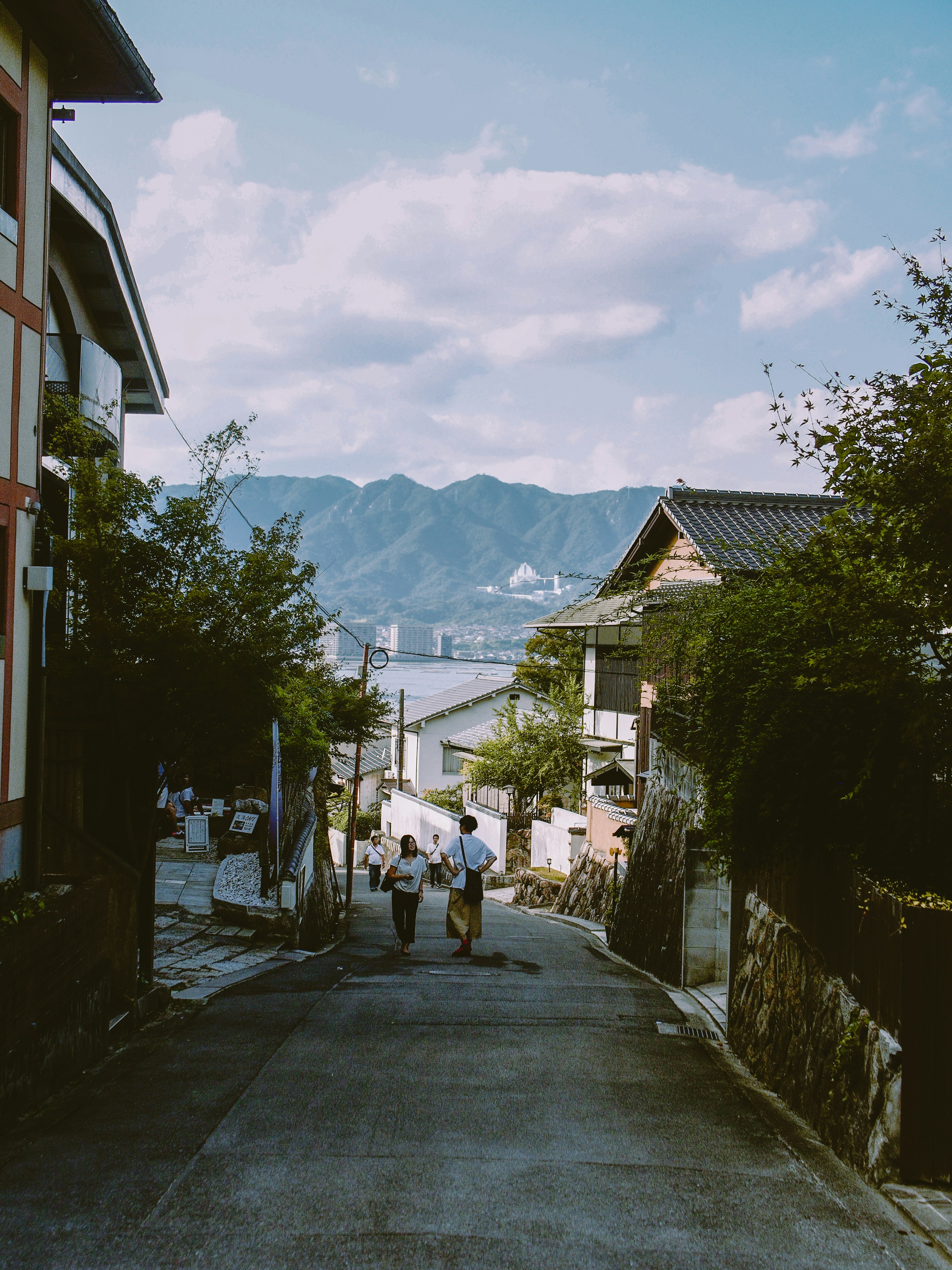Two figures stroll down a tranquil street lined with traditional architecture, framed by distant mountains under a blue sky.