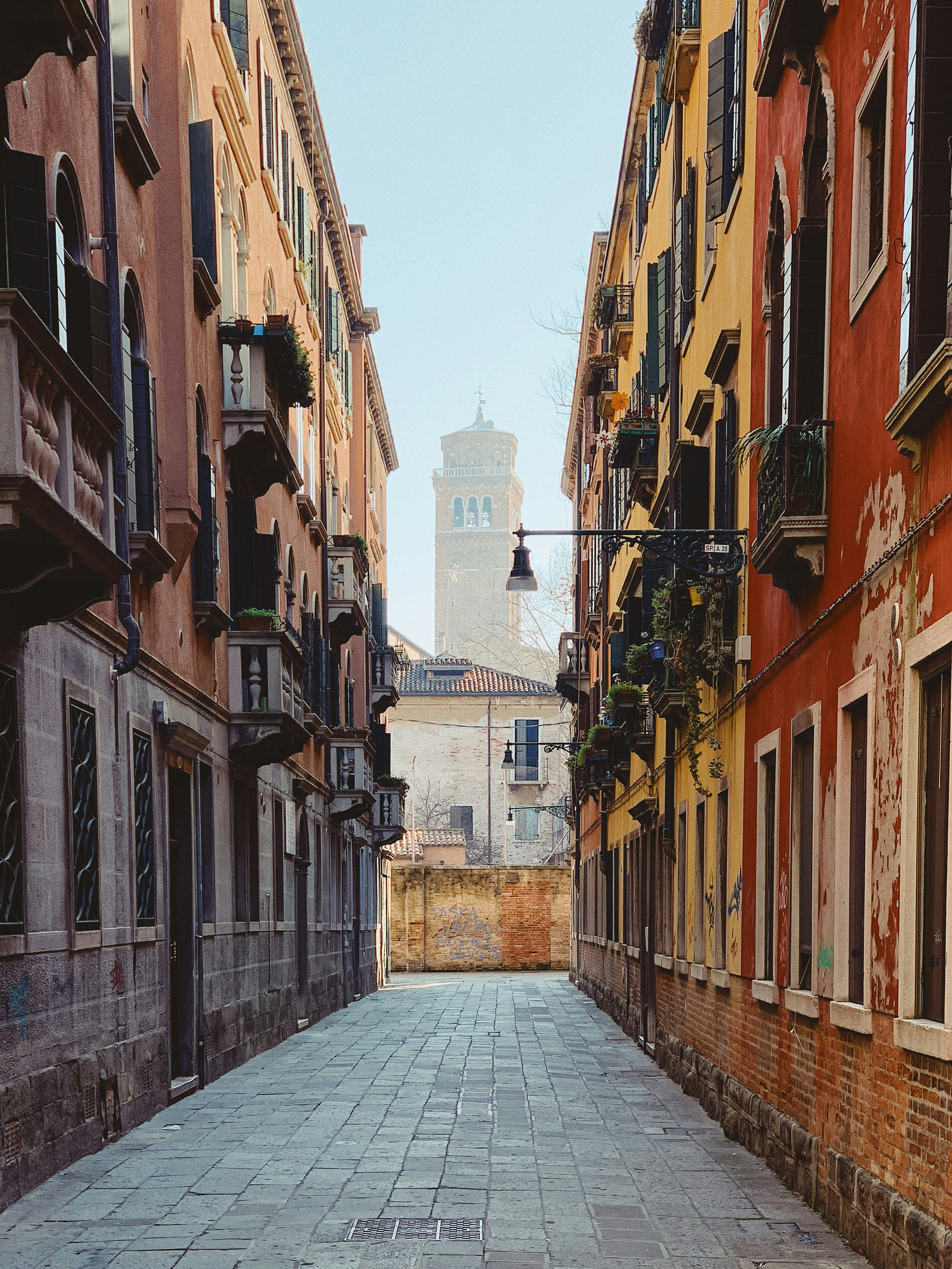 Narrow alleyway in Venice flanked by colorful buildings, leading to a distant bell tower. The scene captures the charm and tranquility of the city.