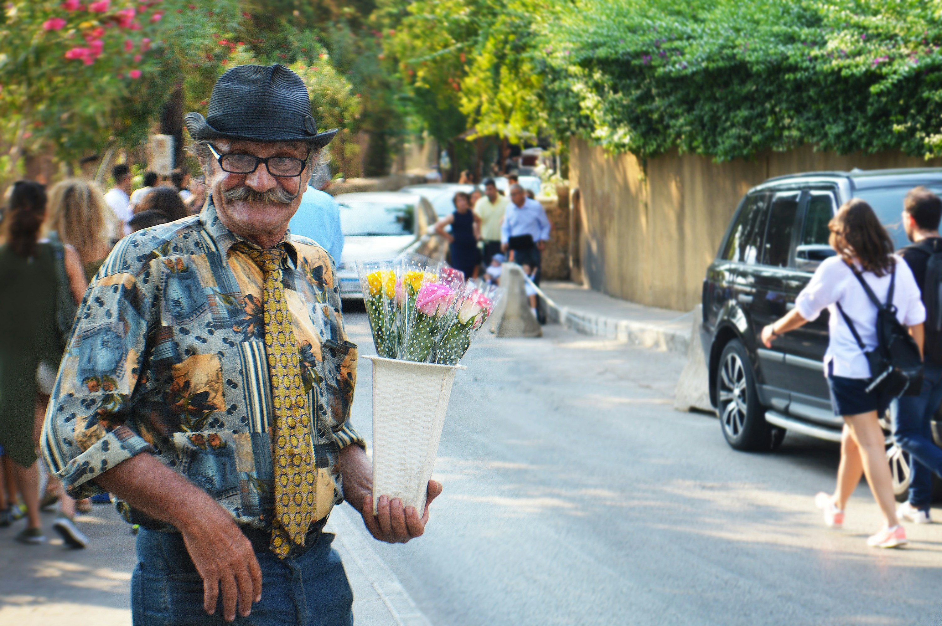 Elderly man in a hat holds a bouquet of flowers on a bustling street, surrounded by pedestrians and cars.
