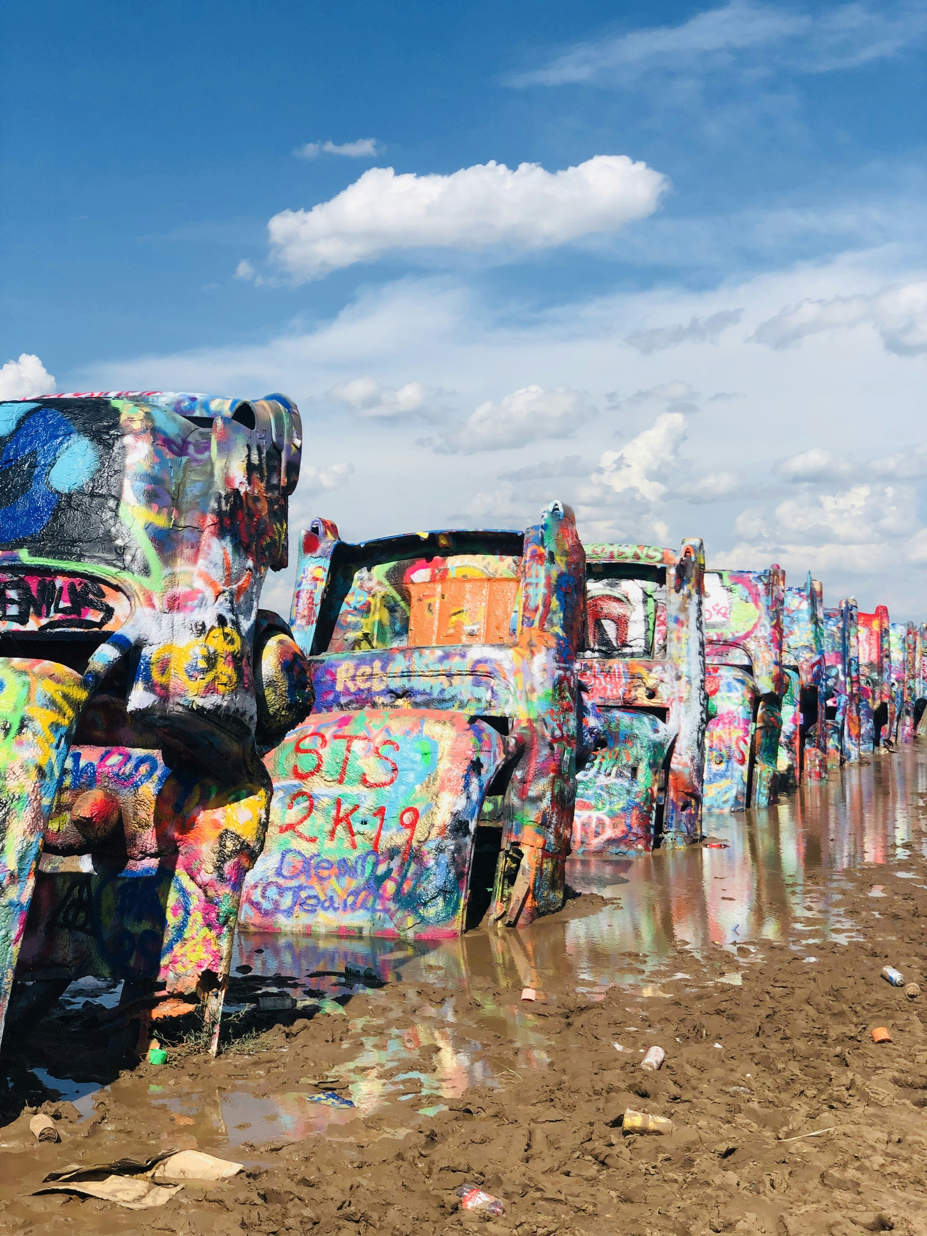 Colorfully painted vintage cars partially submerged in muddy ground under a bright blue sky with fluffy clouds.