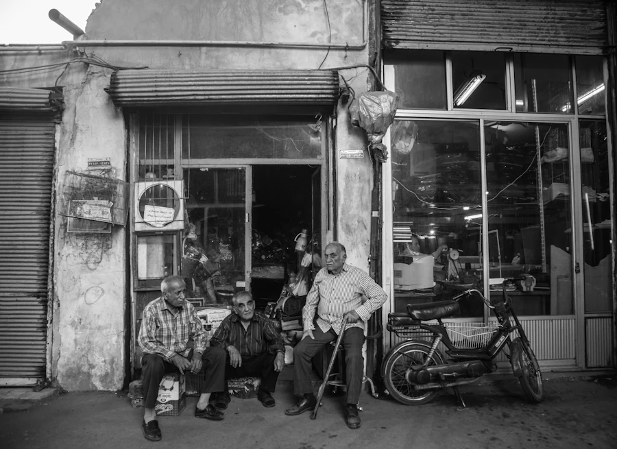 Three older men sit and stand in front of a worn storefront, with a bicycle parked beside them. The scene is rendered in black and white, with reflections in the shop windows and a quiet street mood.