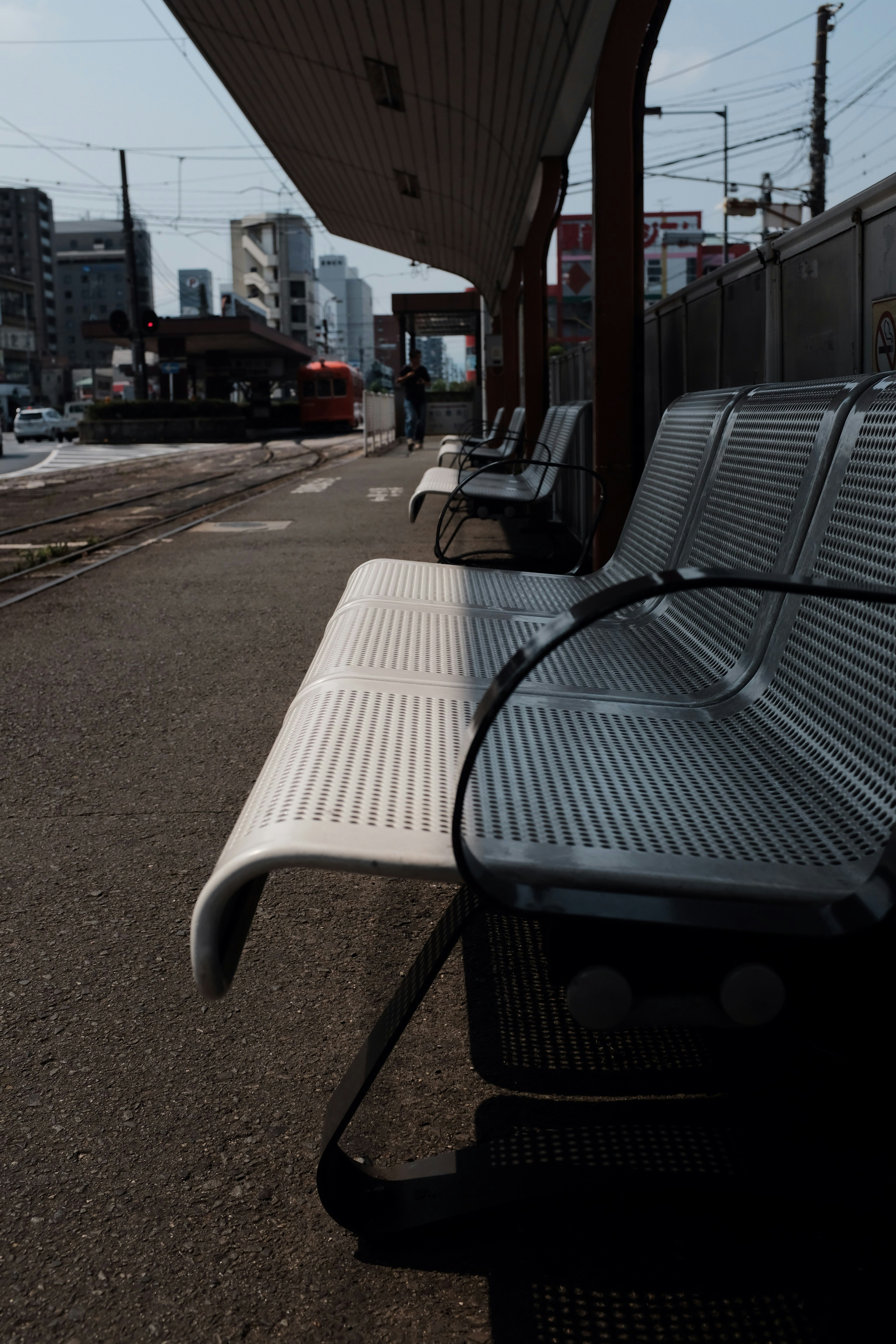Empty metal benches at a city tram station, with a hint of urban life in the background. A red tram is seen in the distance.