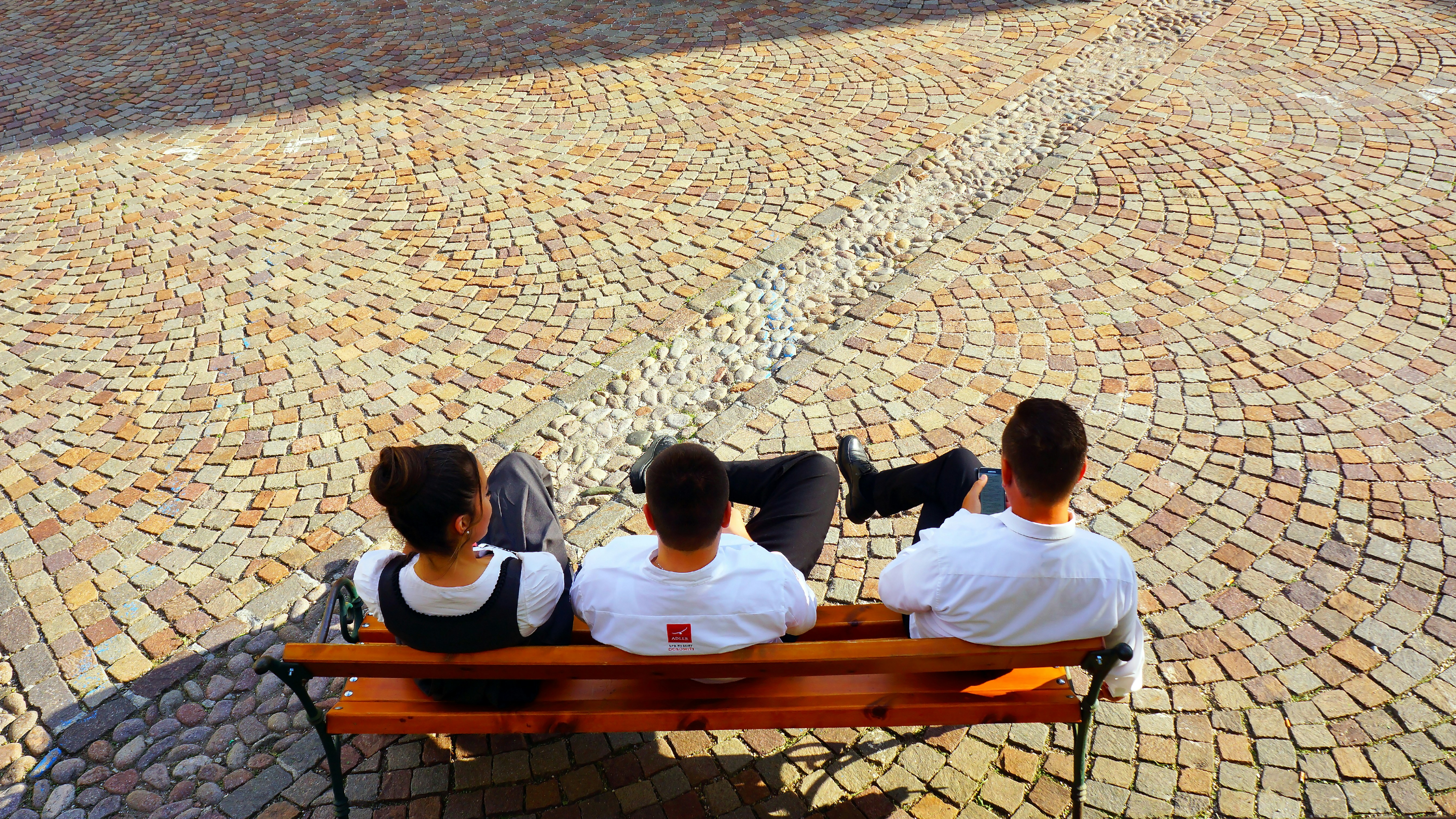Three friends on a sunlit park bench, tiled courtyard stretching outward