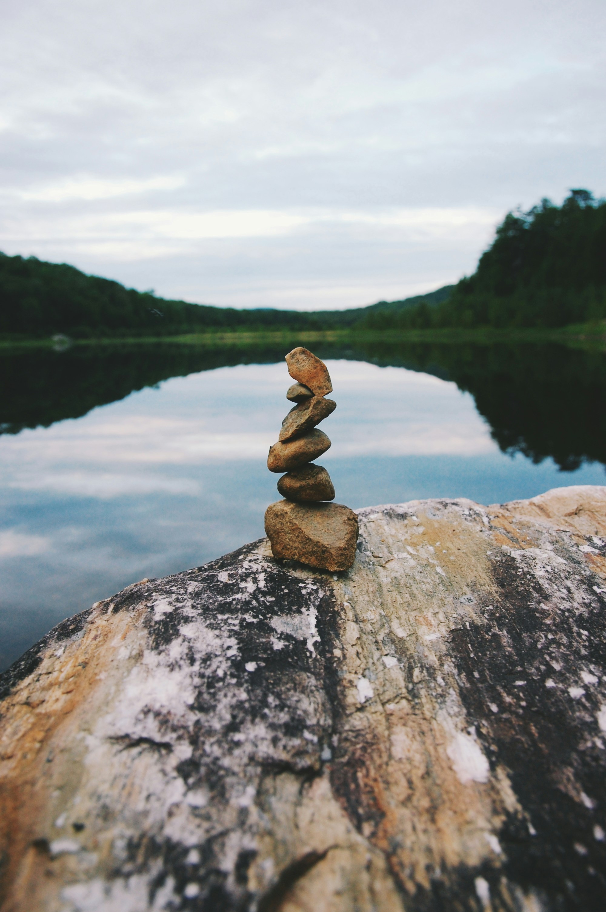 Stacked stones on a log by a tranquil lake, with forested hills in the background under a cloudy sky.