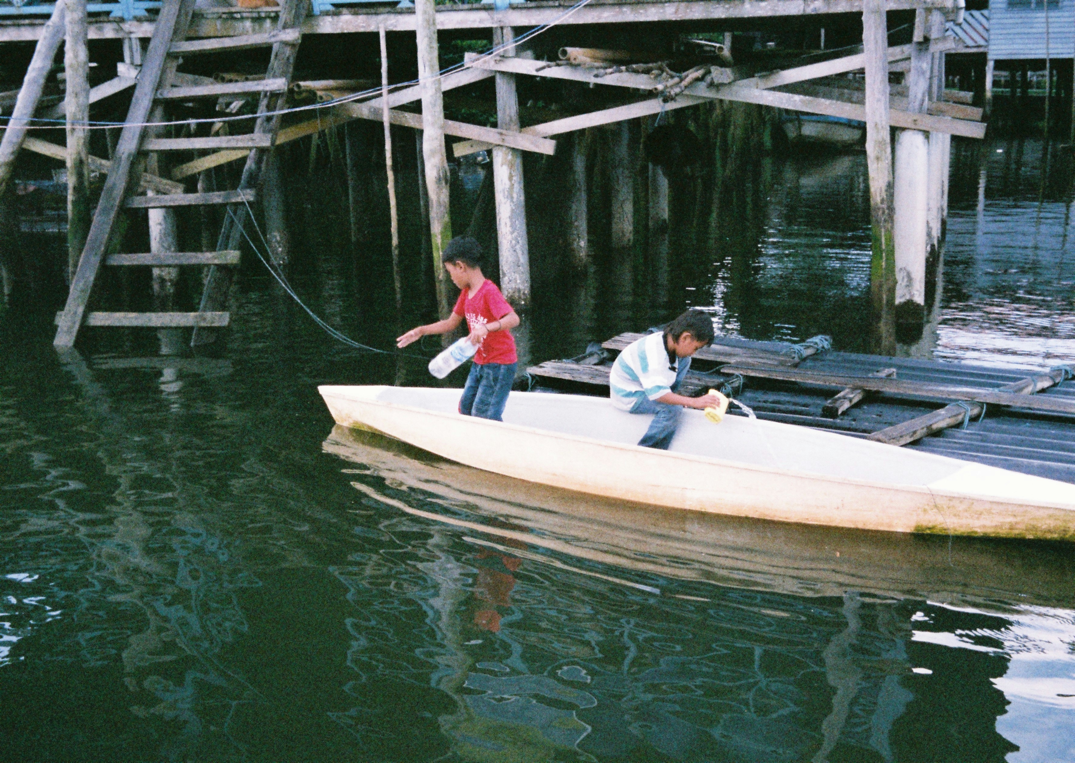 Two children fishing from a small boat near a wooden pier, surrounded by calm waters reflecting their playful activity.