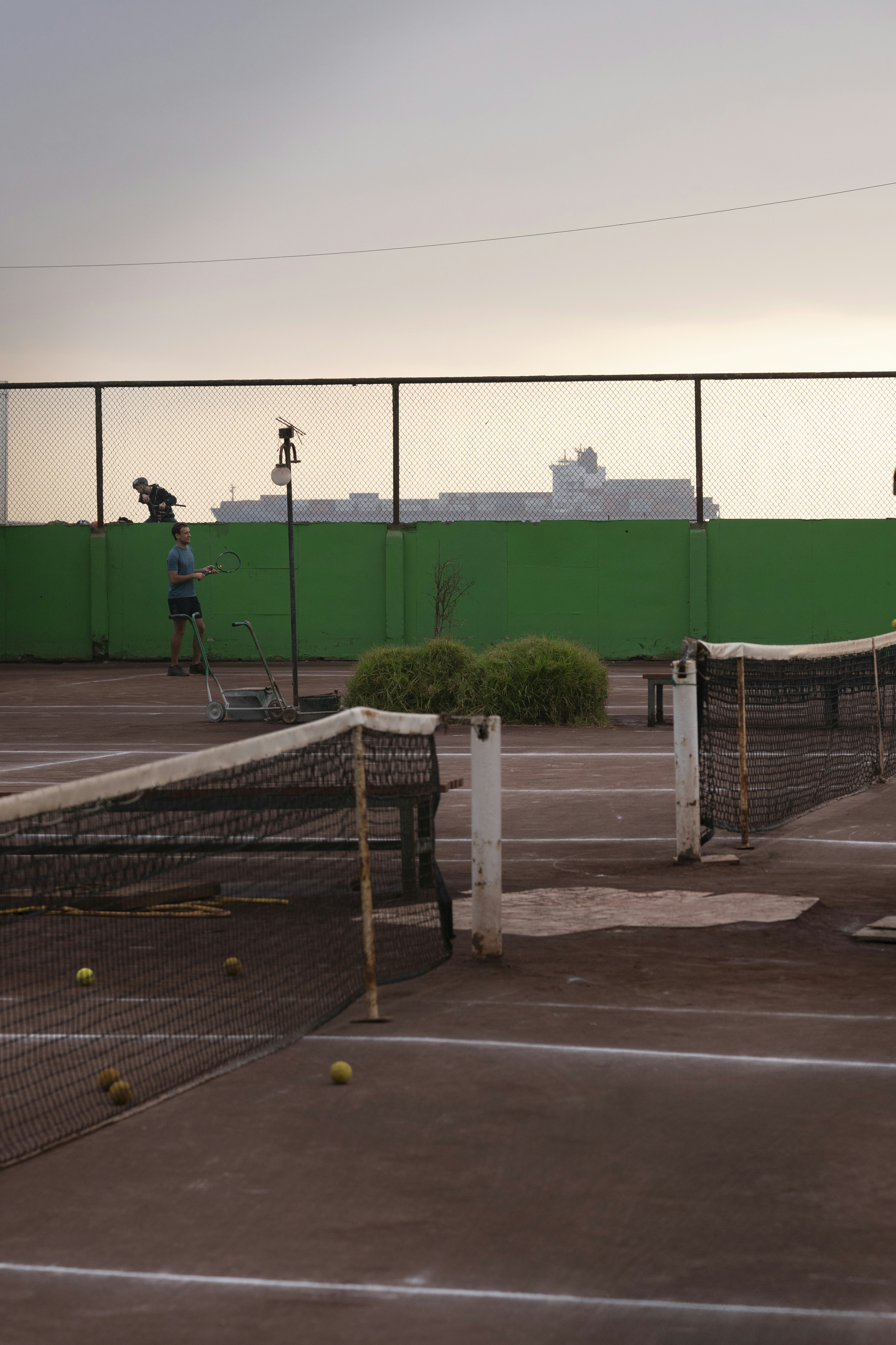 Tennis courts at dusk with a lone player and camera gear behind a green barrier; soft light and long shadows define the empty foreground.