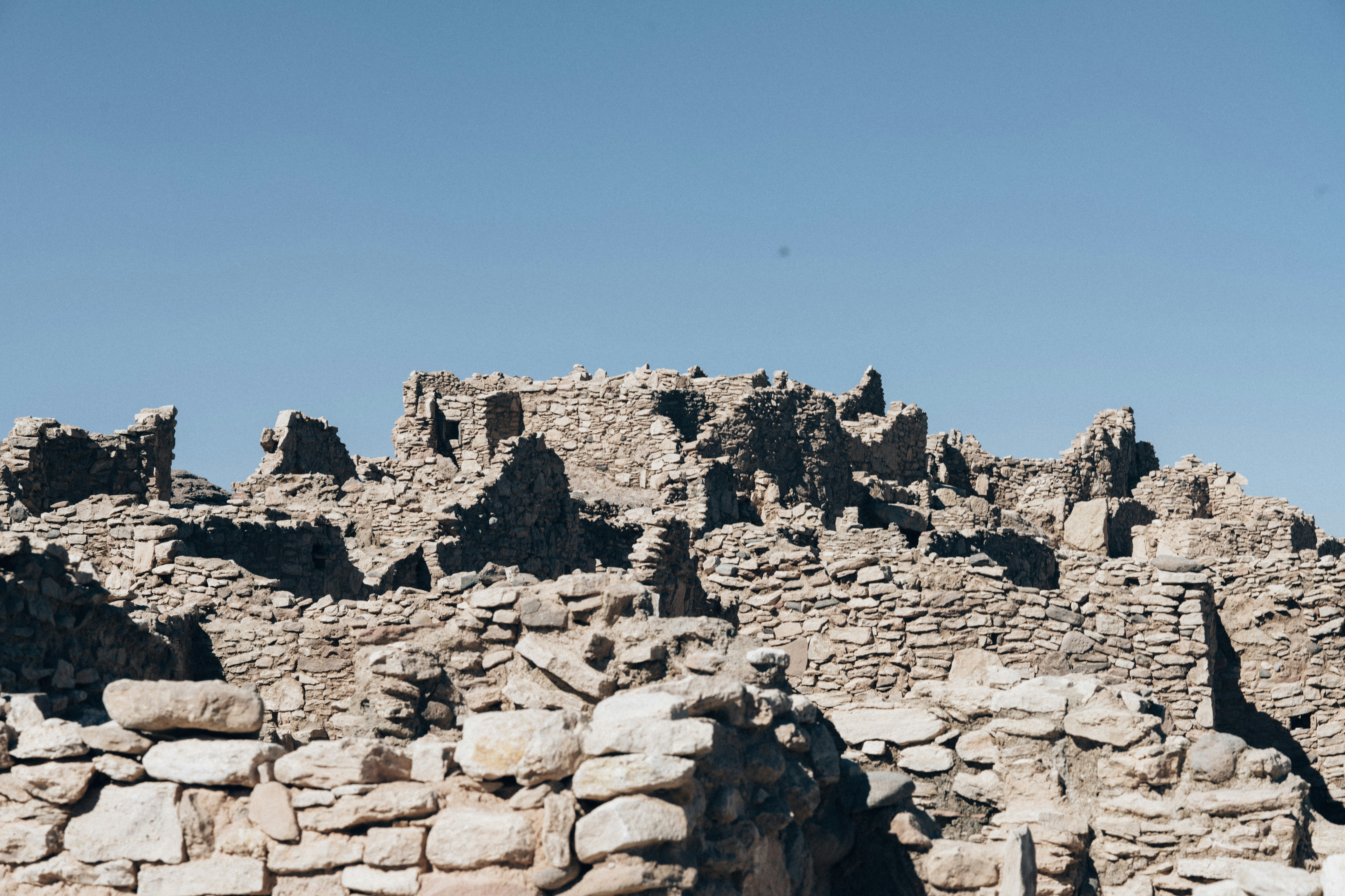 Weathered stone ruins of an ancient settlement against a clear blue sky. The remnants showcase intricate stonework and historical significance.