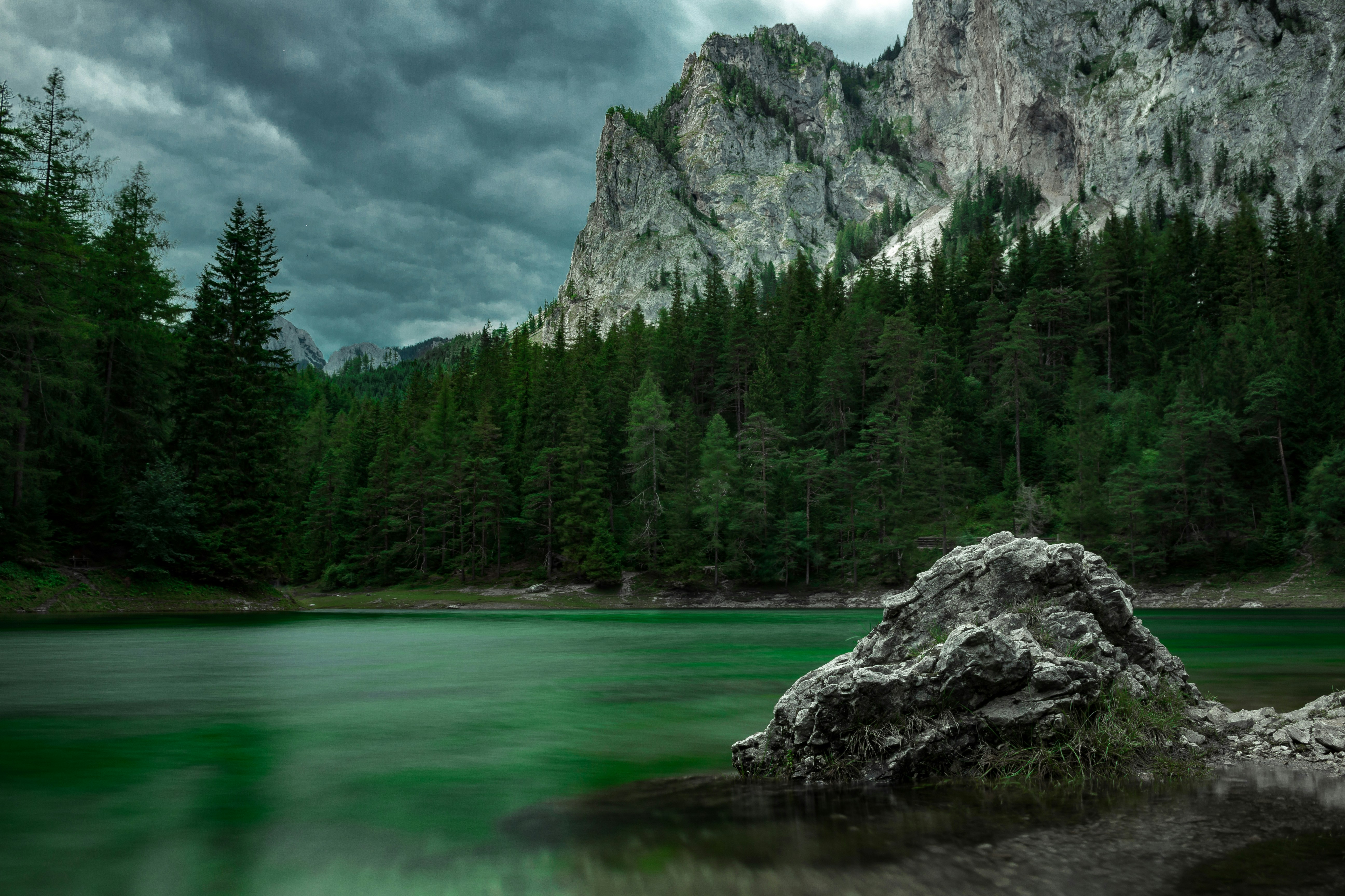 A serene lake framed by towering mountains and dense evergreen trees, with a prominent rock in the foreground. The moody sky hints at an impending storm.