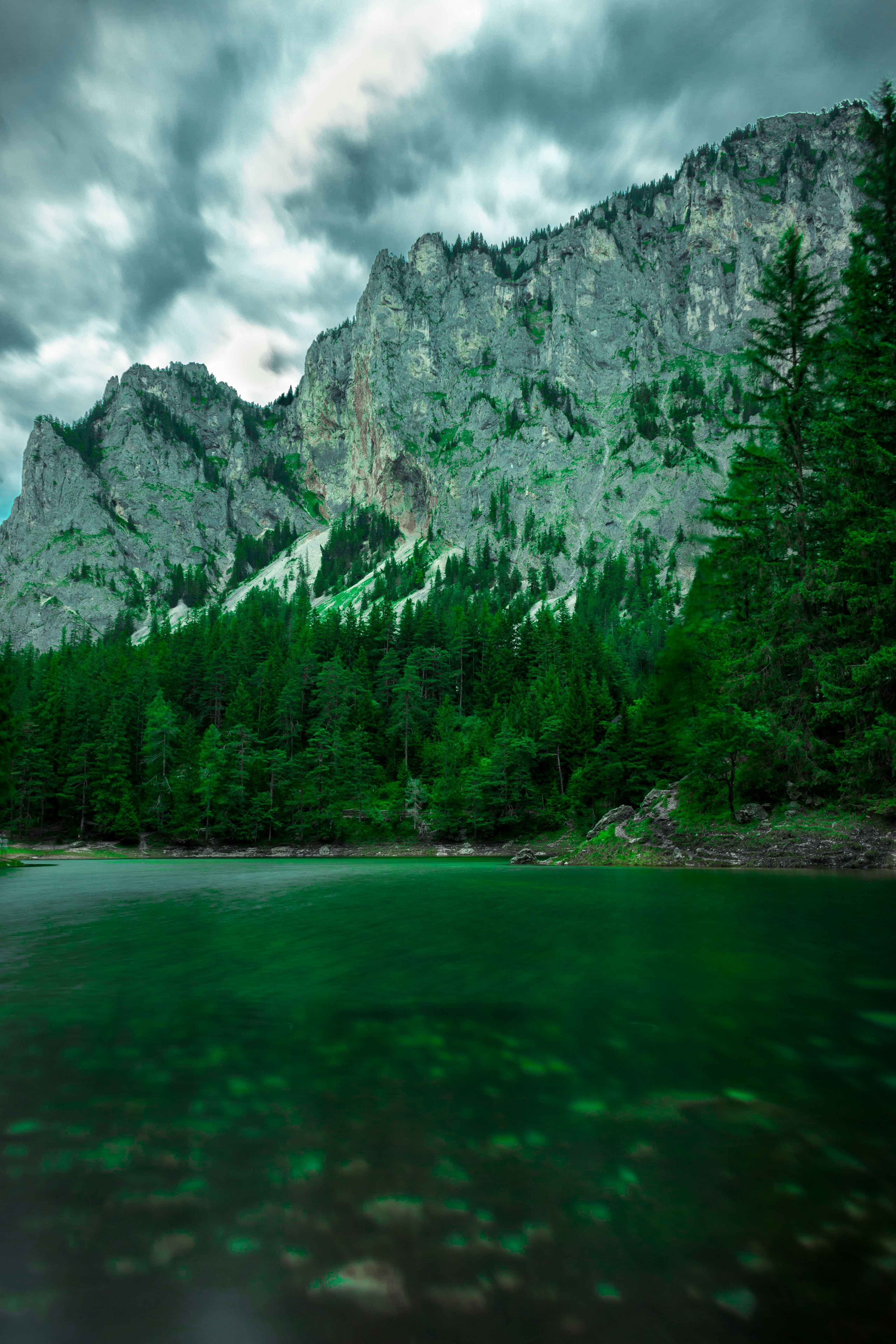 Majestic mountain range looming over a serene, emerald-green lake surrounded by lush coniferous trees.