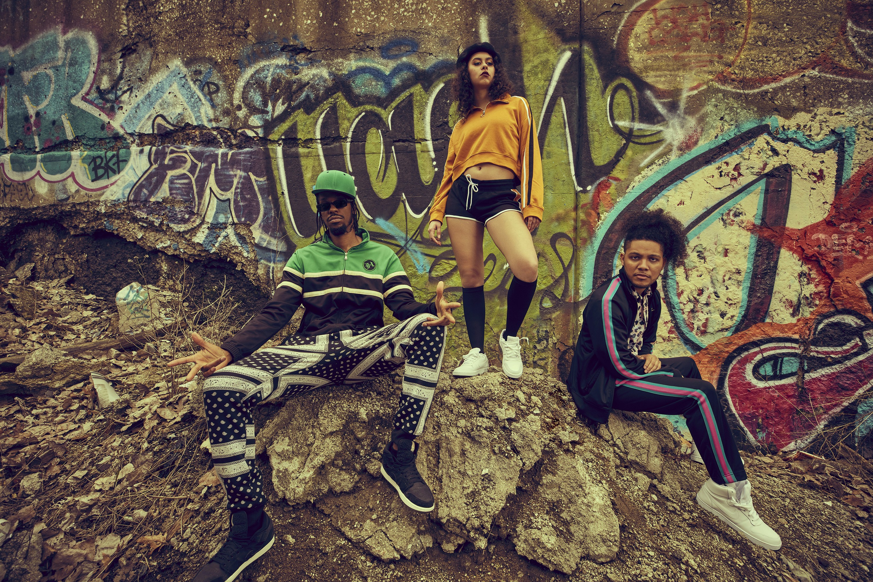 Three individuals posing against a vibrant graffiti backdrop, showcasing contemporary fashion and urban culture.