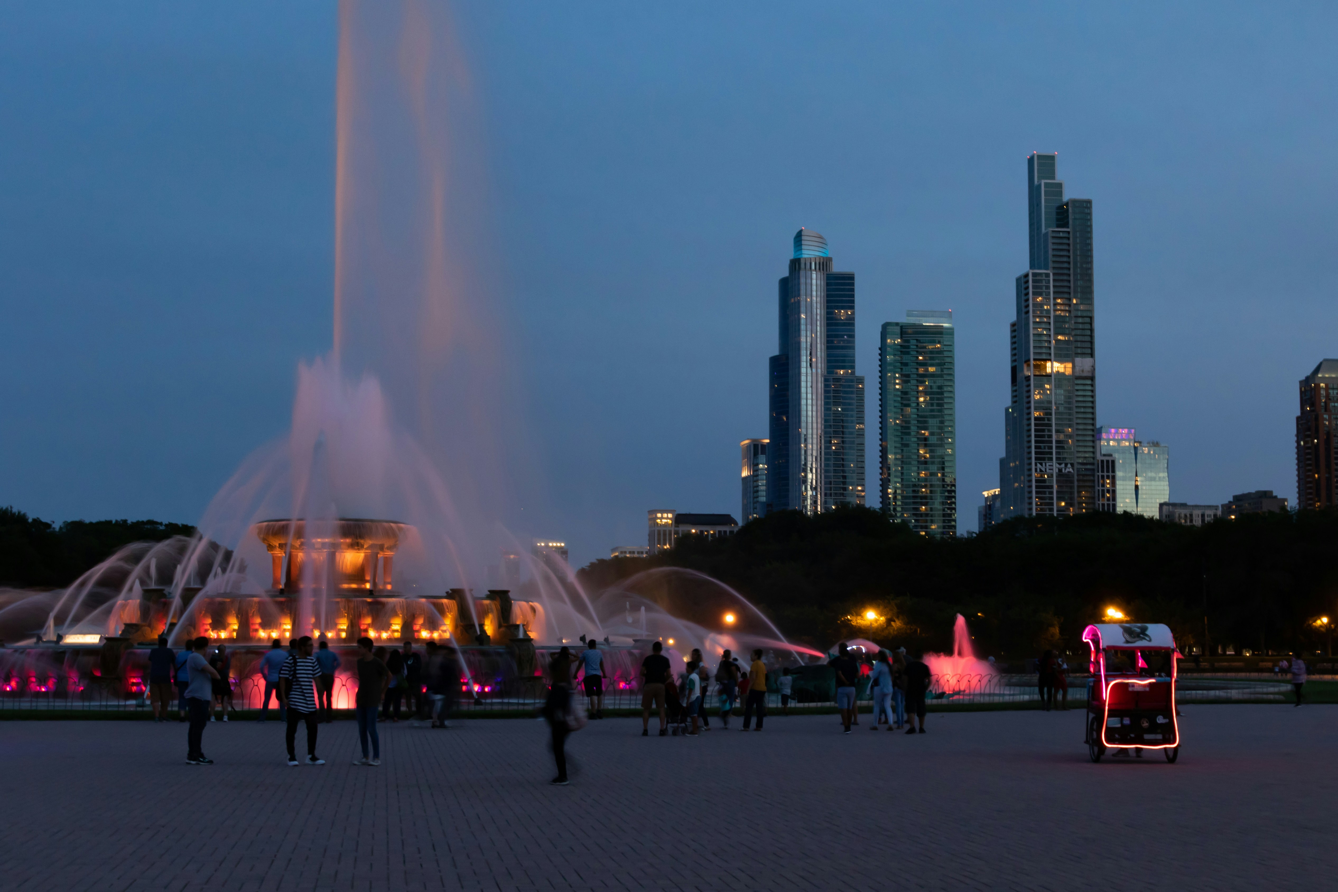 People gather near a brightly lit fountain with skyscrapers illuminated in the background.