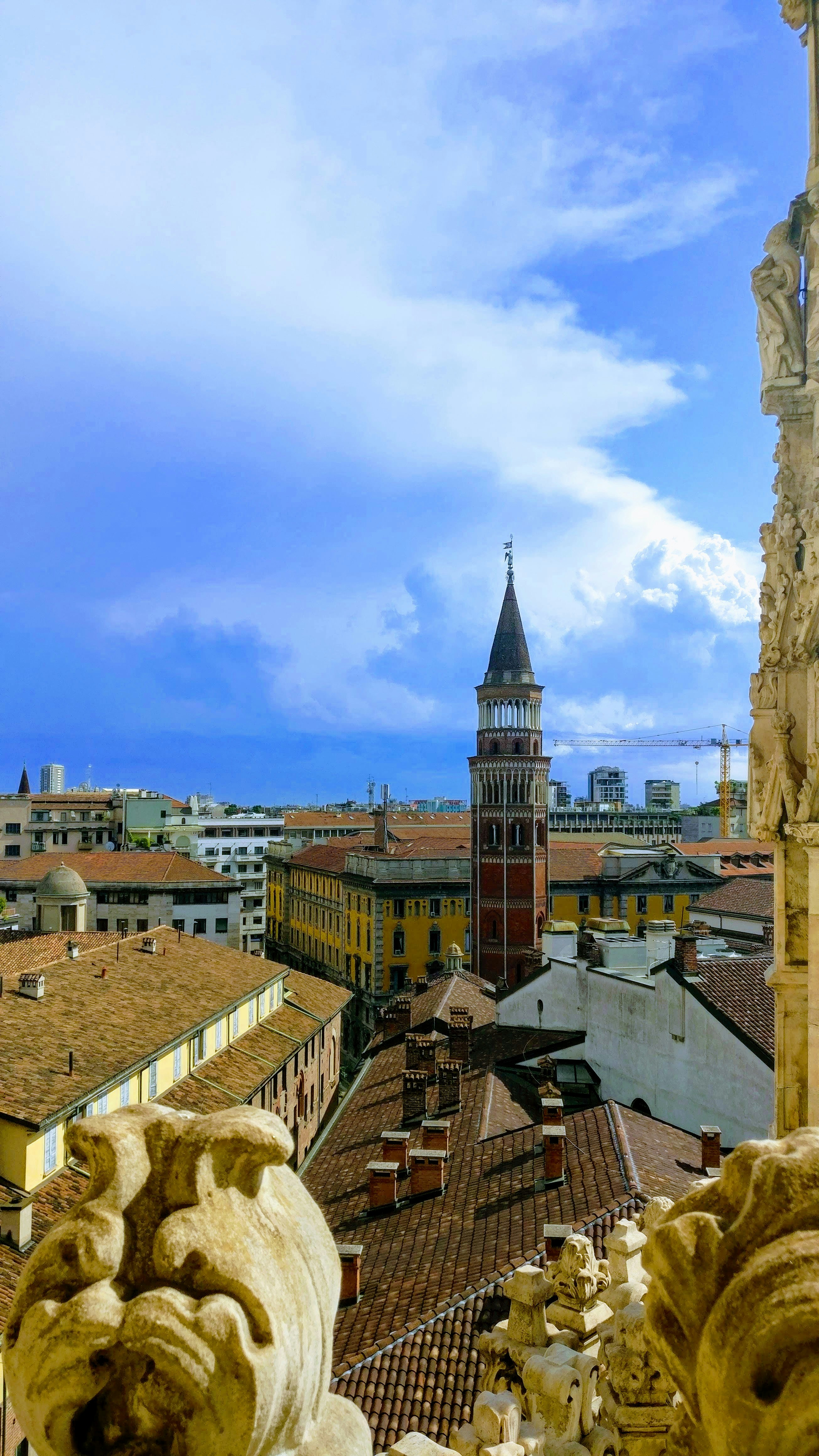 Rooftop cityscape with terracotta tiles and ornate stone balustrades, foreground framing a tall brick campanile against a bright blue sky.