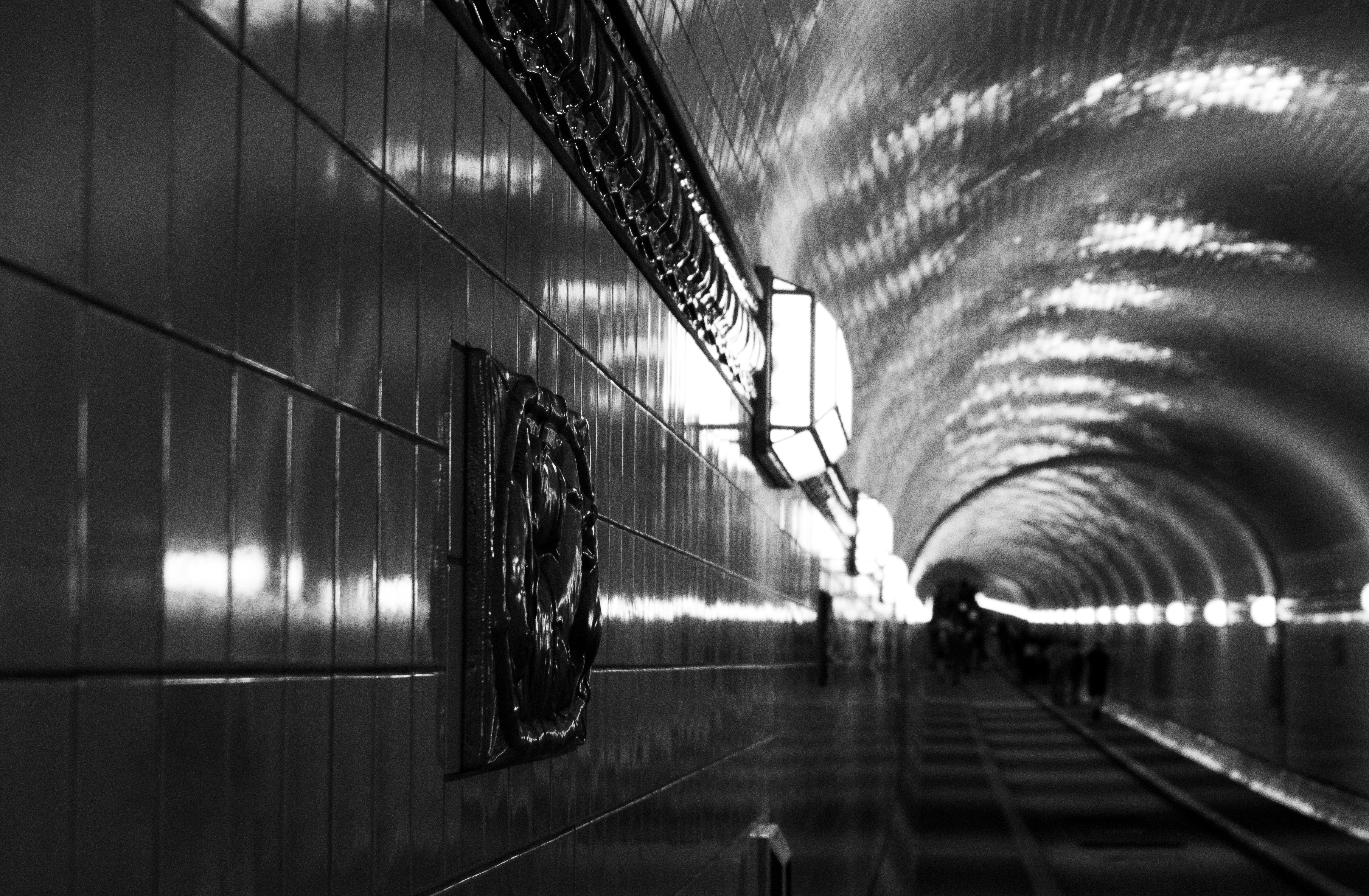 Grayscale subway hallway with intricate wall tiles and glowing lamps creating rhythmic patterns along the tunnel.